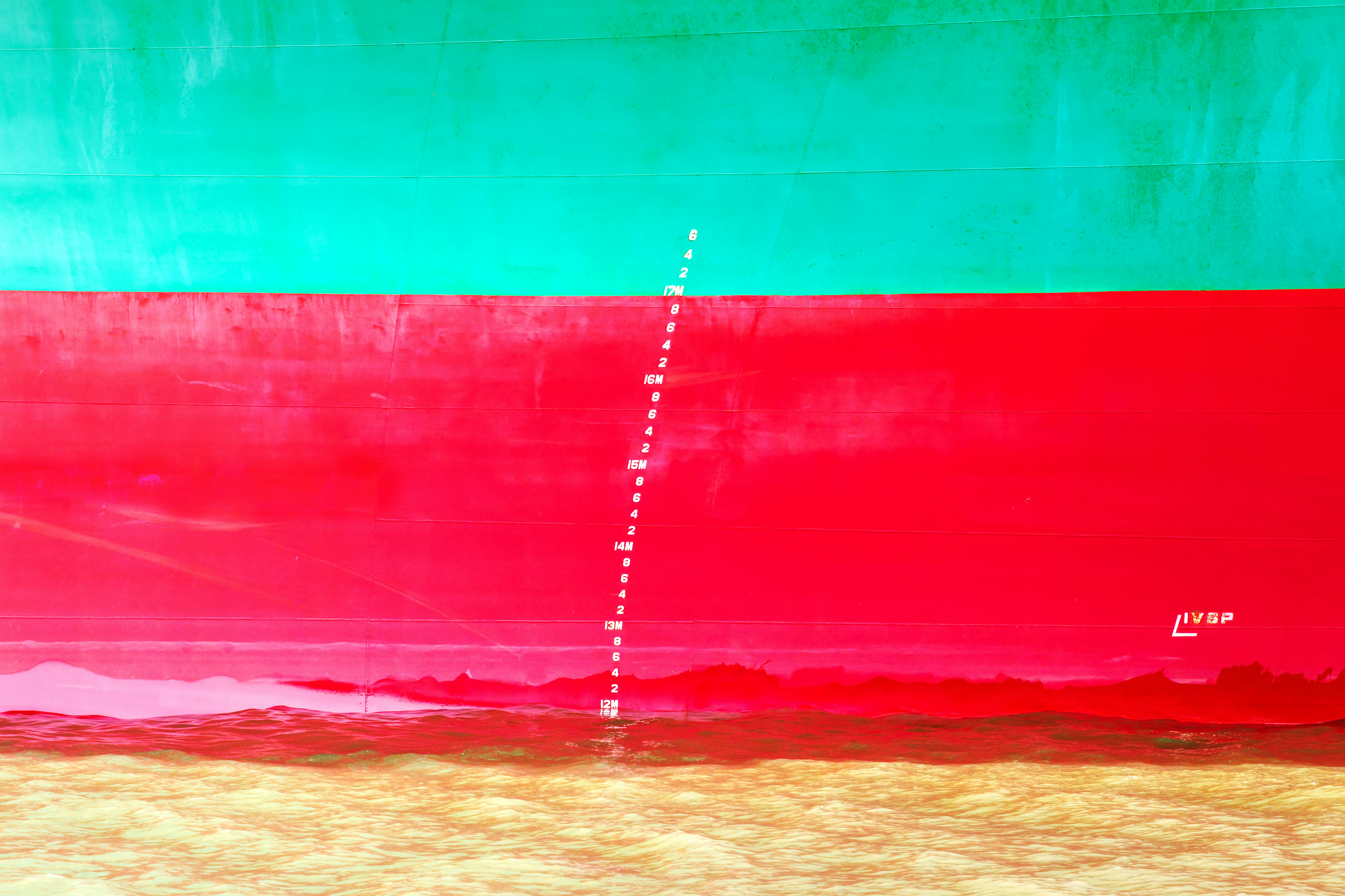 Photograph of a color-blocked wall with teal on top and red below, intersected by a diagonal dotted white line. The sandy foreground adds texture and depth.