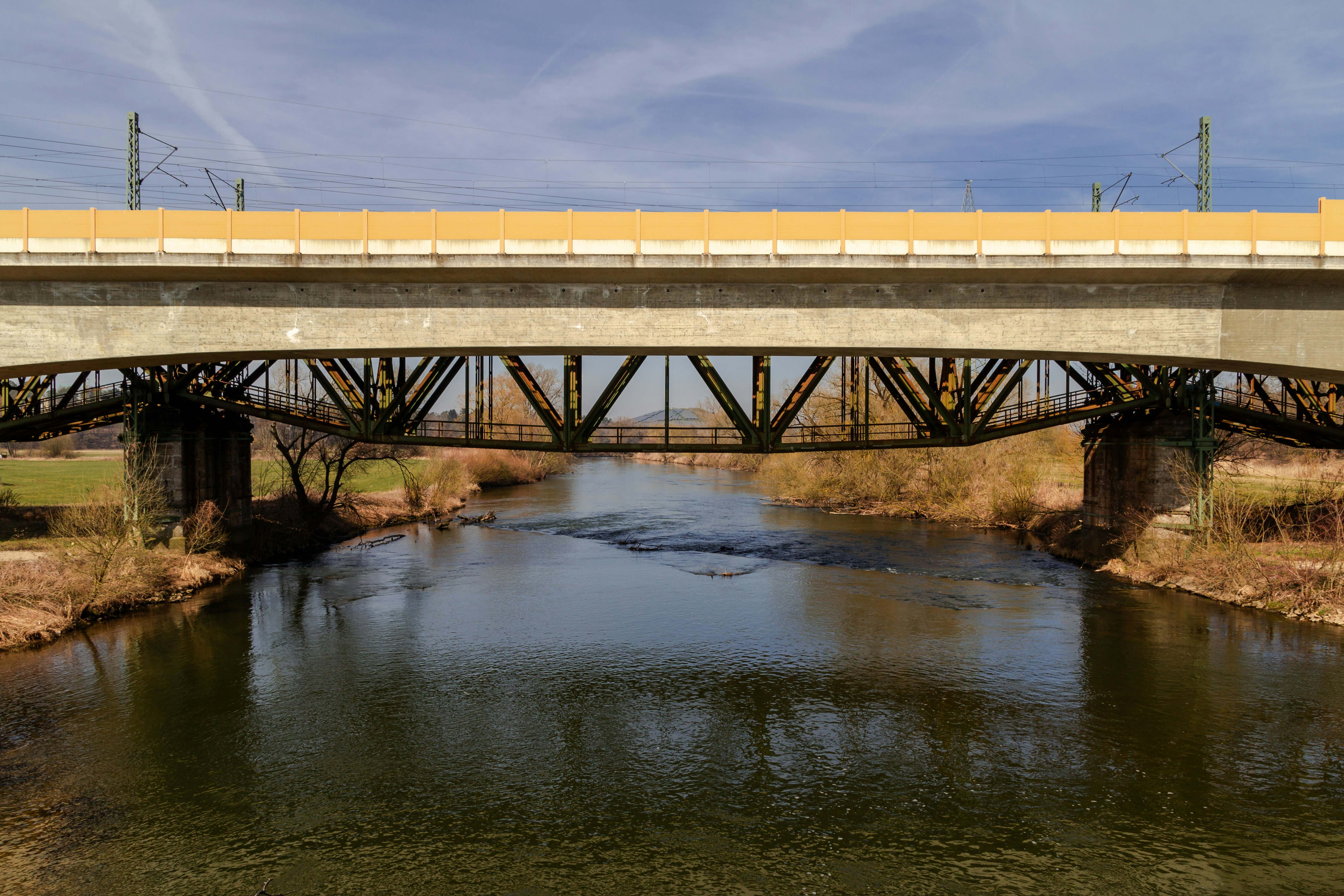 Concrete bridge with metal railings spans a calm river, reflecting the clear blue sky and sparse trees along the banks.