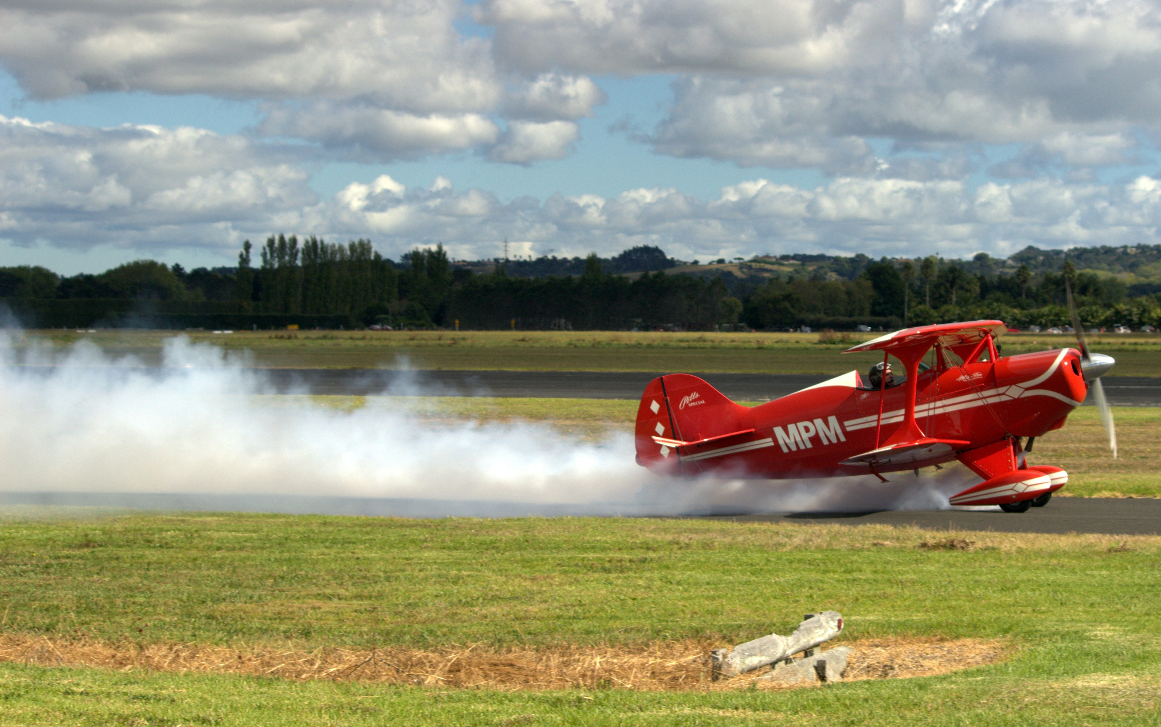 Red biplane performing a smoke-filled maneuver on a runway under a partly cloudy sky.