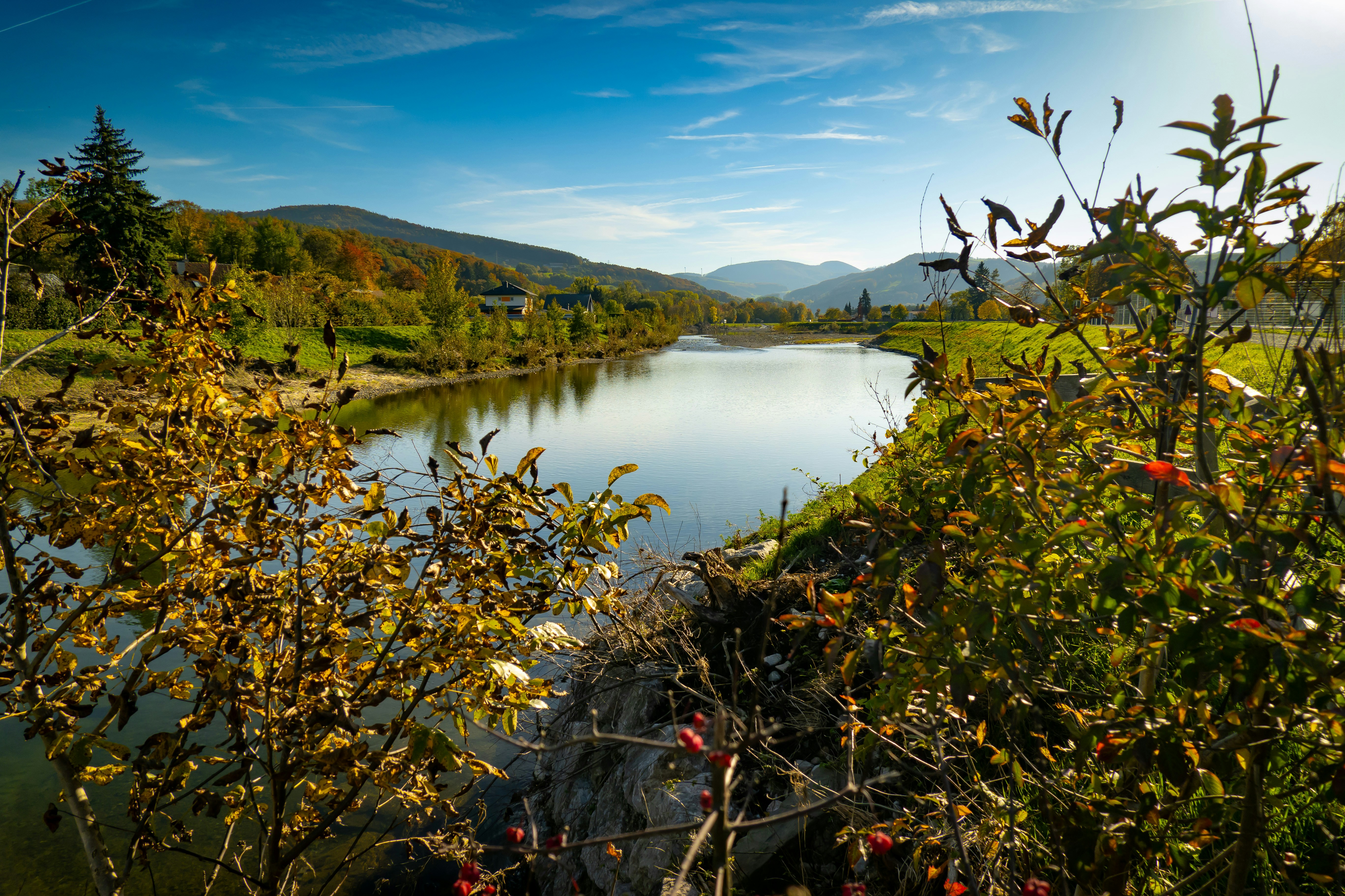 Serene river flanked by autumn foliage and distant mountains under a clear blue sky.