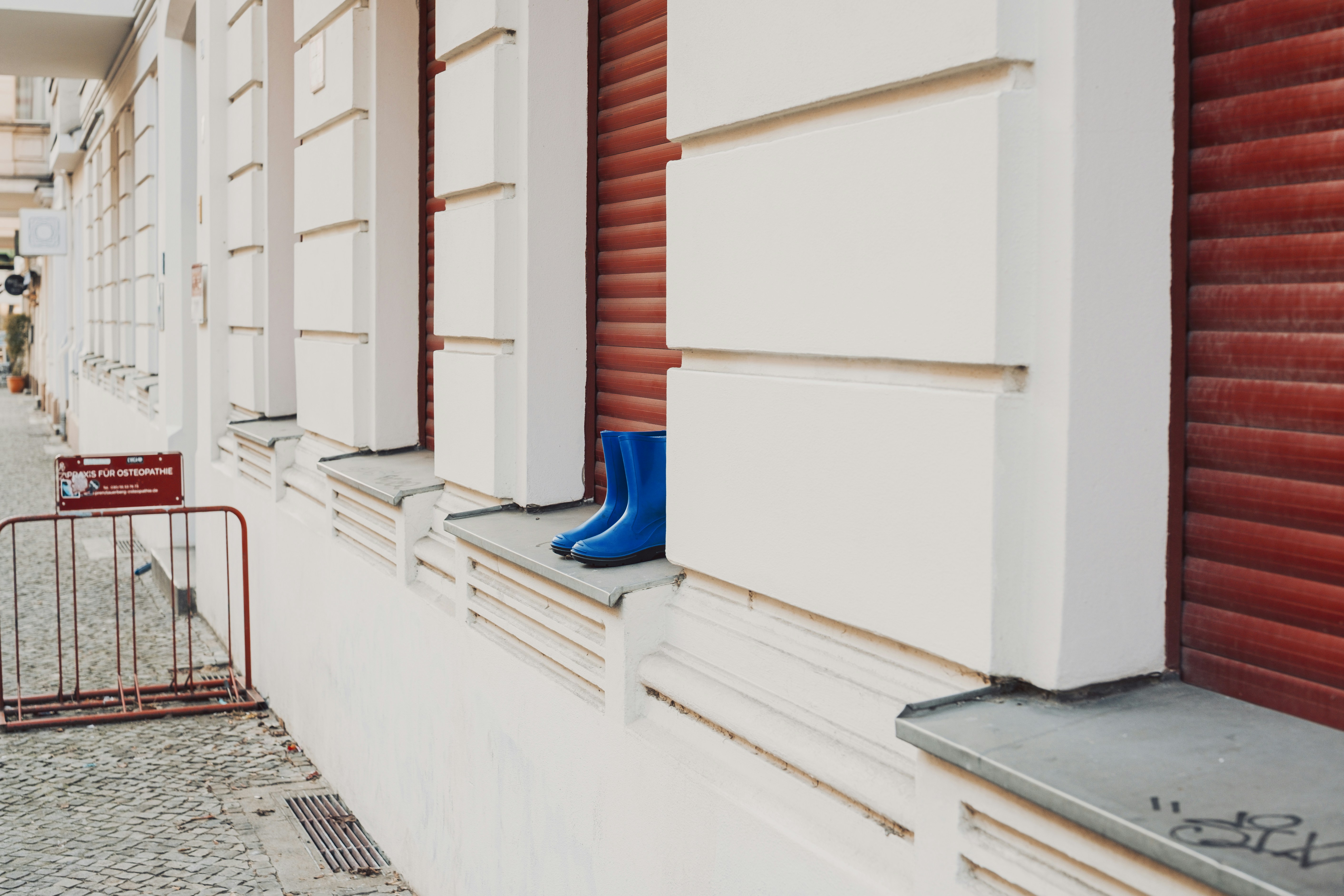 Blue boots resting on a windowsill beside red shutters and white building columns.