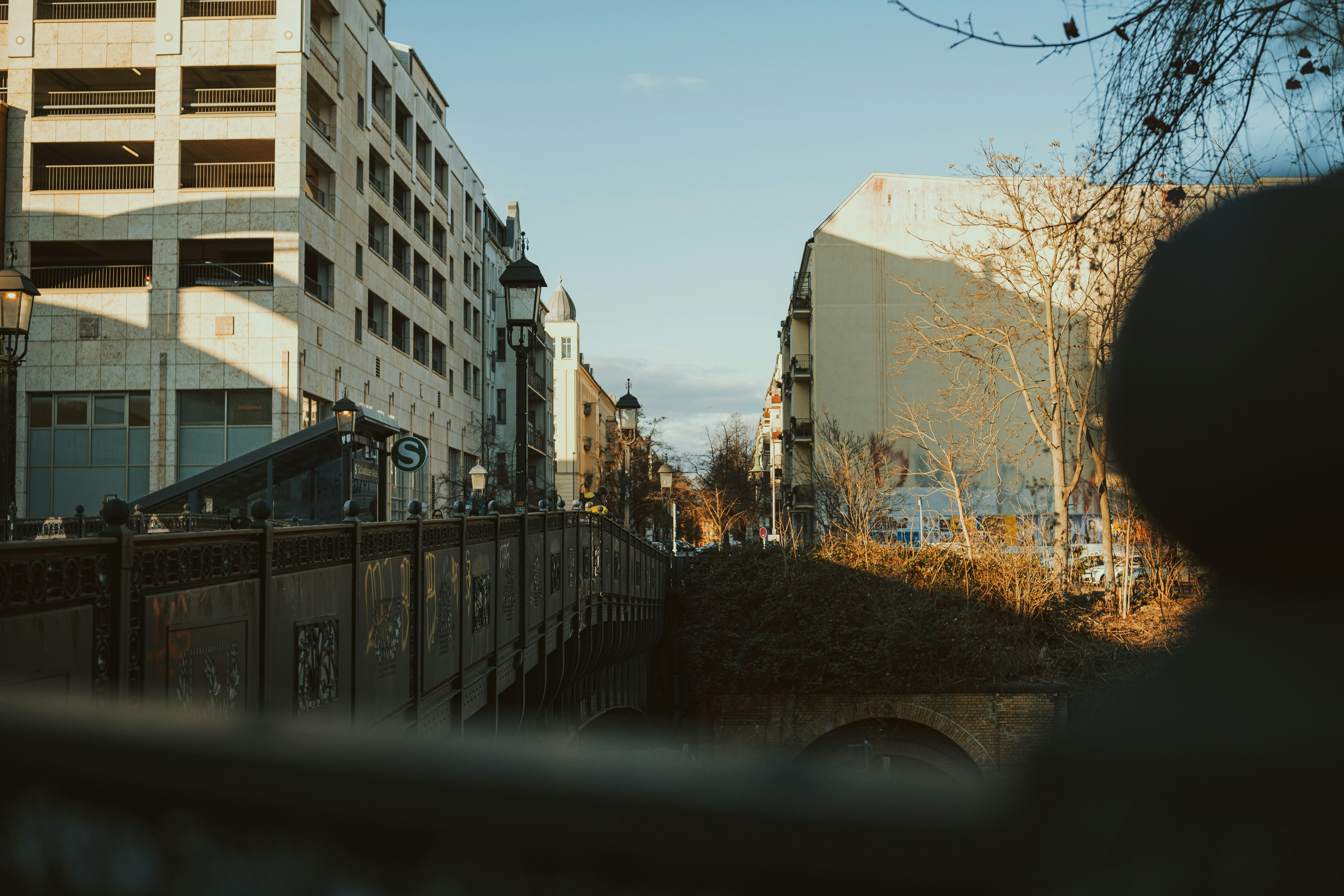 Bridge lined with ornate railings leads towards buildings under a clear sky, bathed in warm sunlight.
