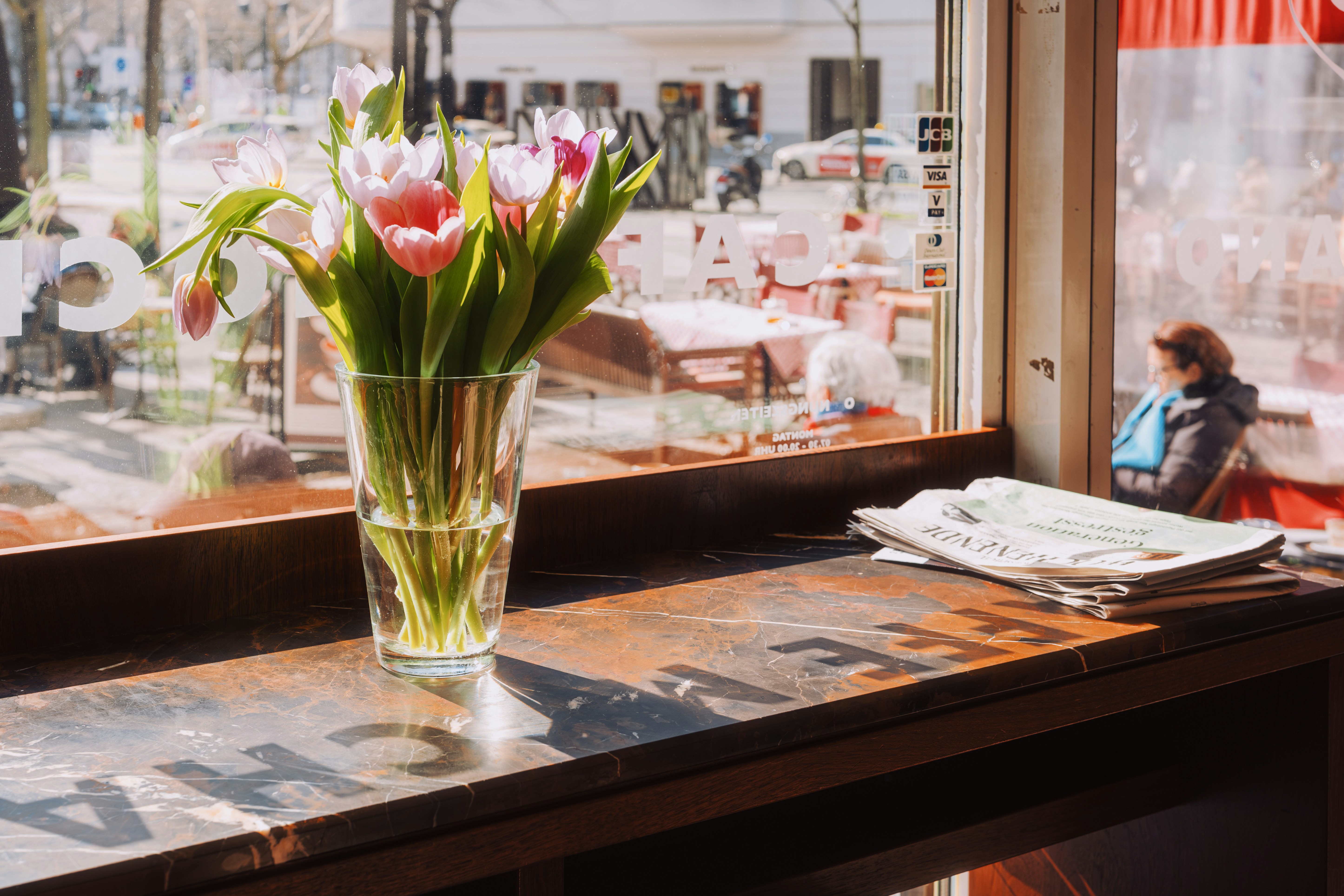Vibrant tulips in a glass vase catch sunlight on a café table, with blurred street scene outside.