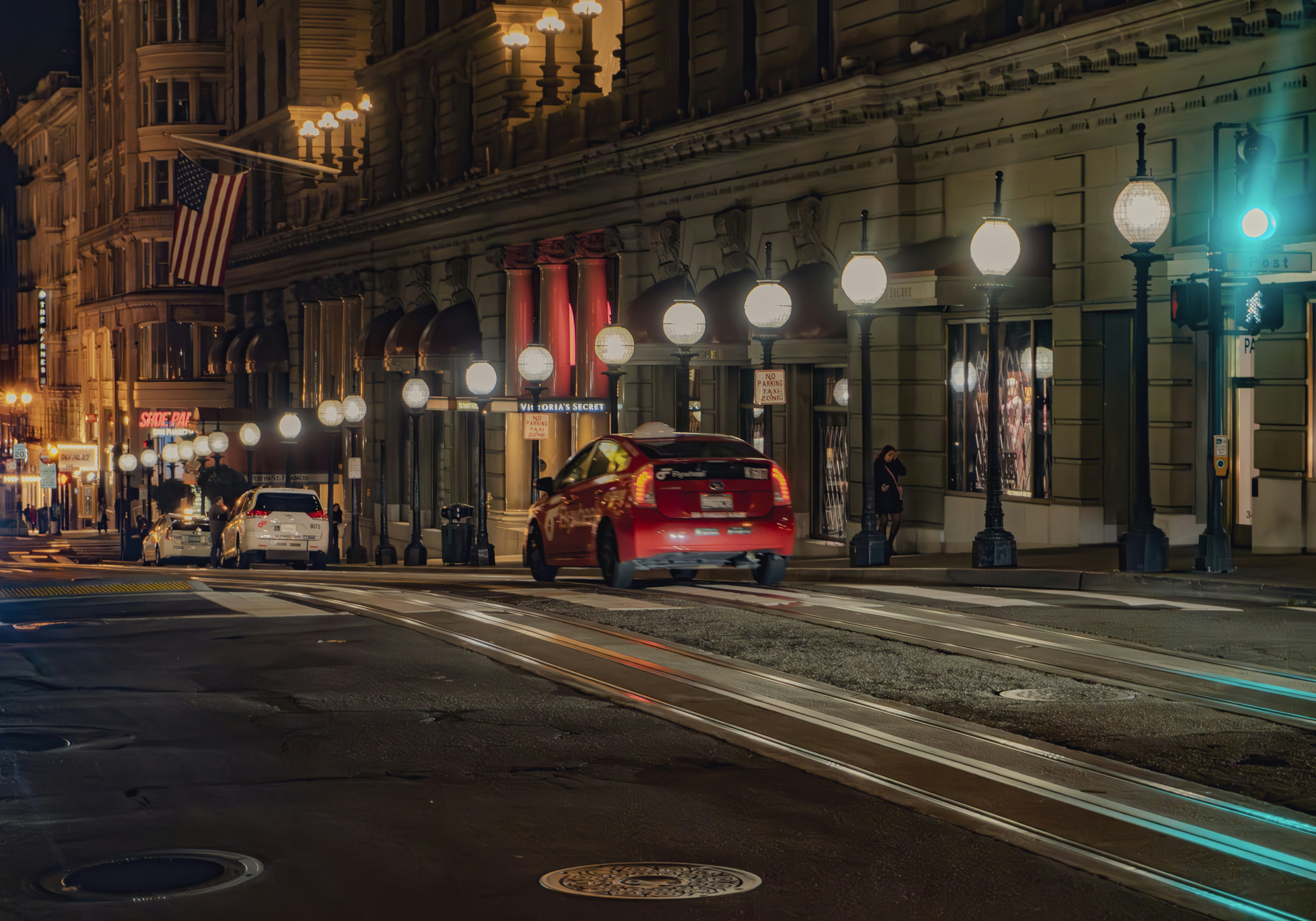 Red car navigating a dimly lit historic street lined with glowing lampposts and flags at night.