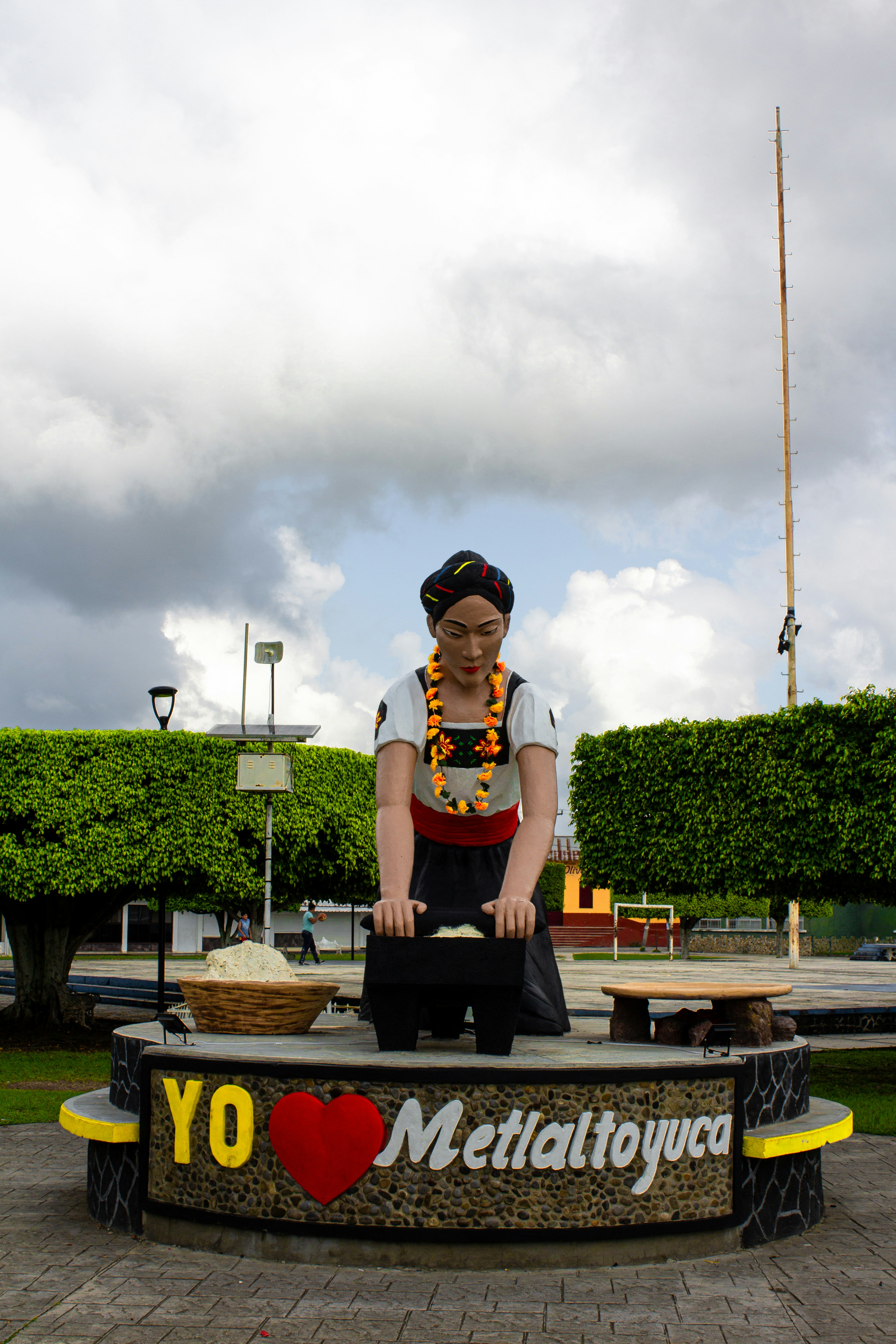 Statue of a traditional figure in festive attire kneading dough in an urban park setting with cloudy skies.