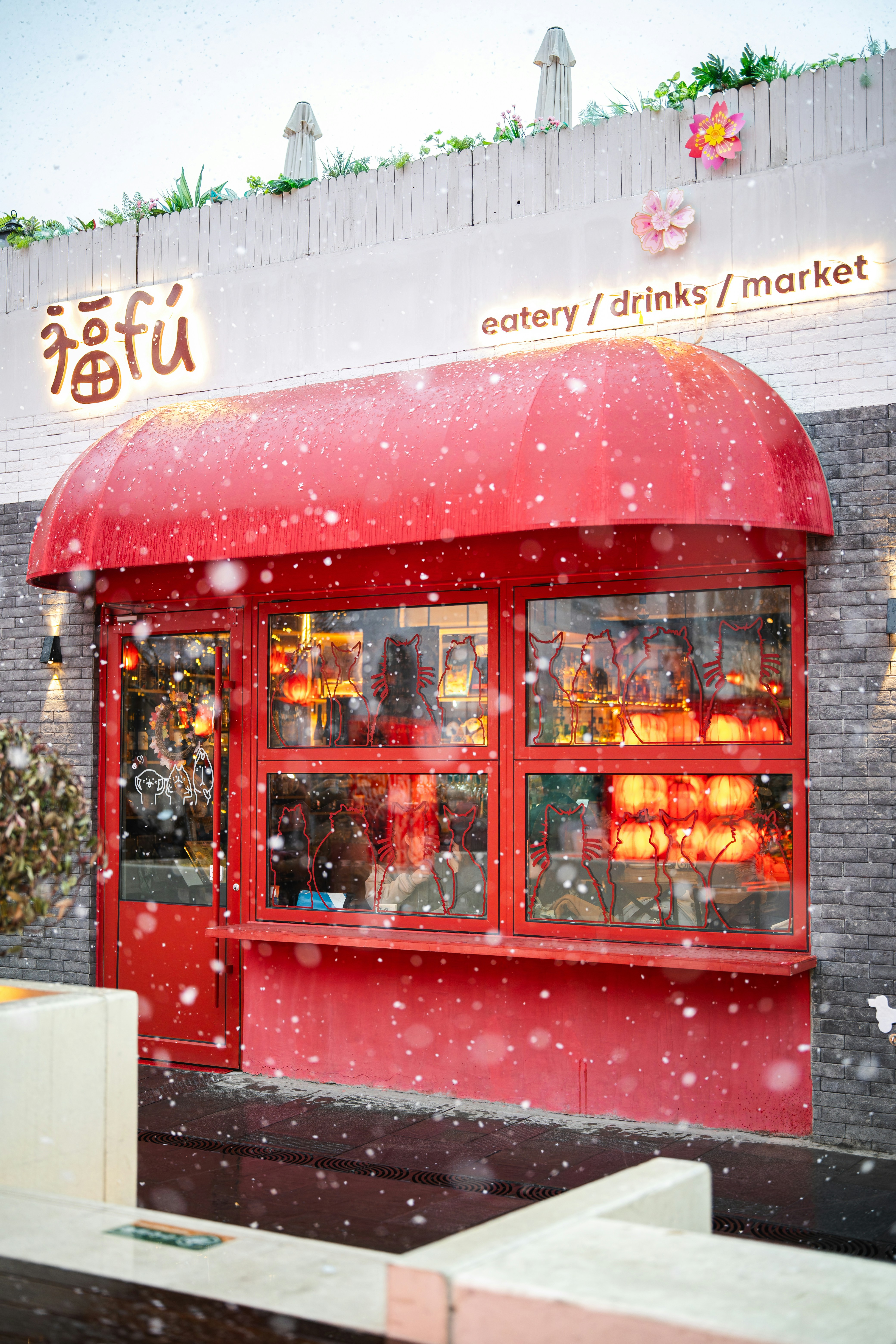 Snowfall outside a cafe with a vibrant red awning and glowing interior lights.