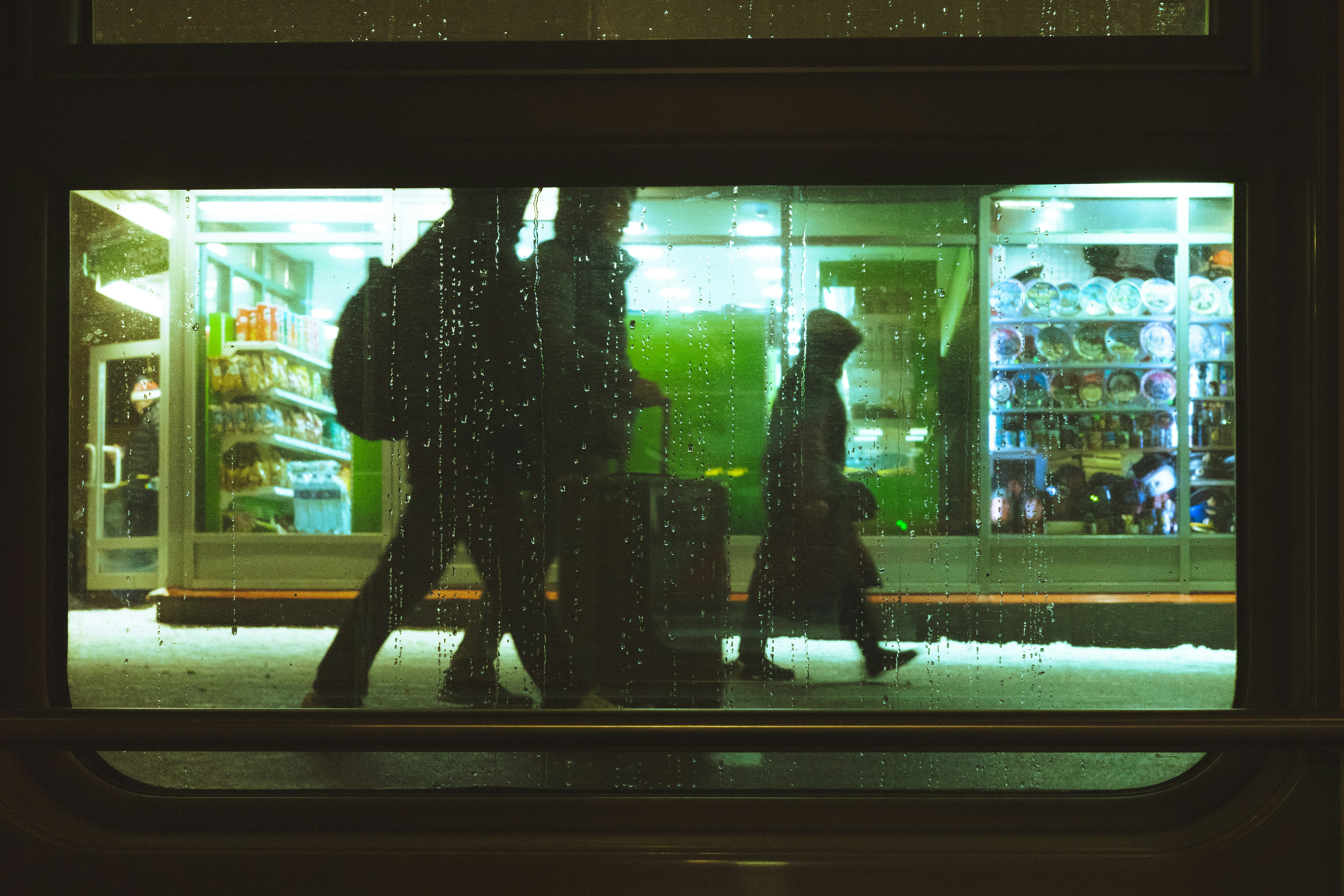 Silhouetted figures walk past an illuminated storefront as seen through a rain-speckled window.