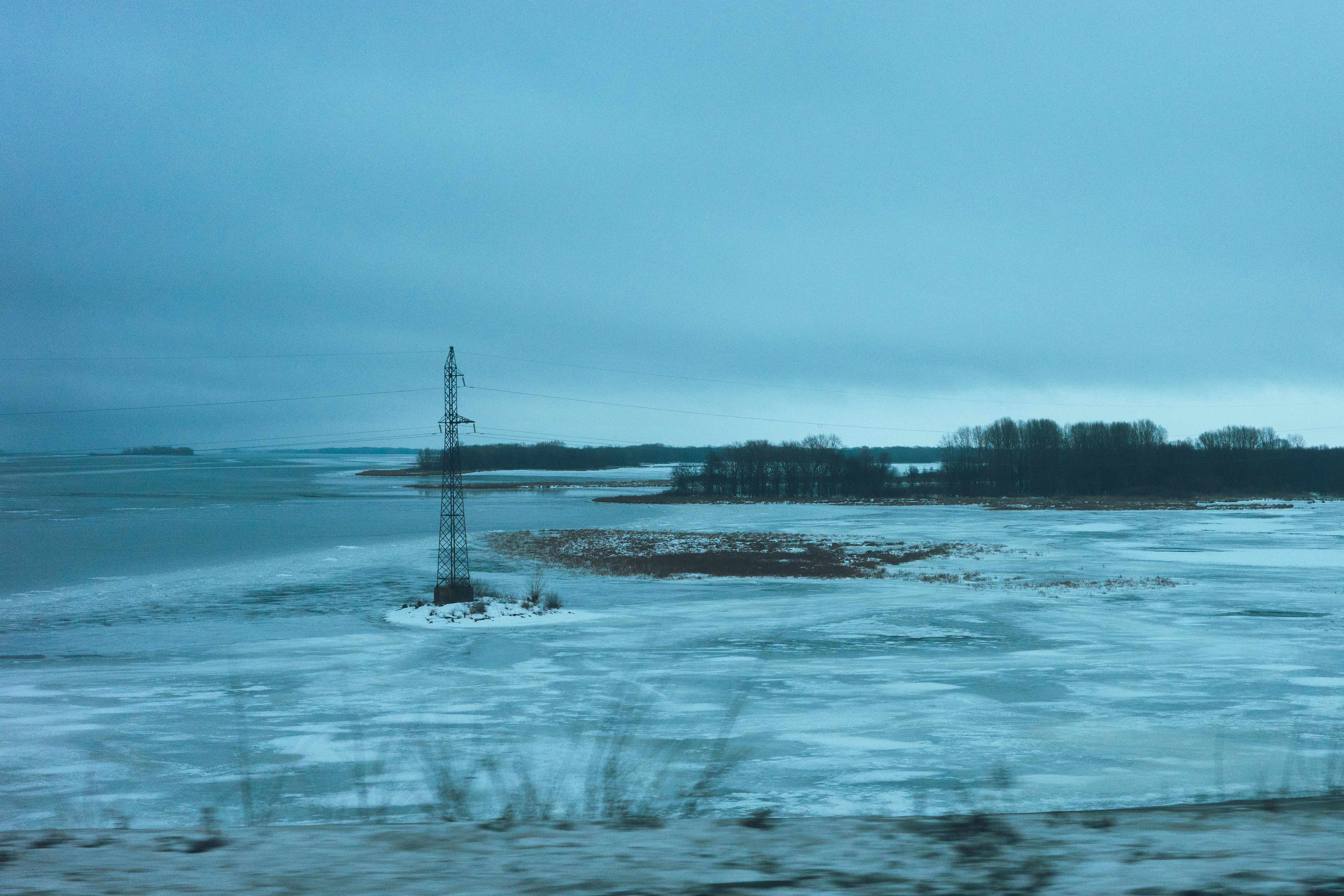 A lone power line tower stands amidst a frozen landscape, surrounded by icy waters and distant trees under a moody sky.