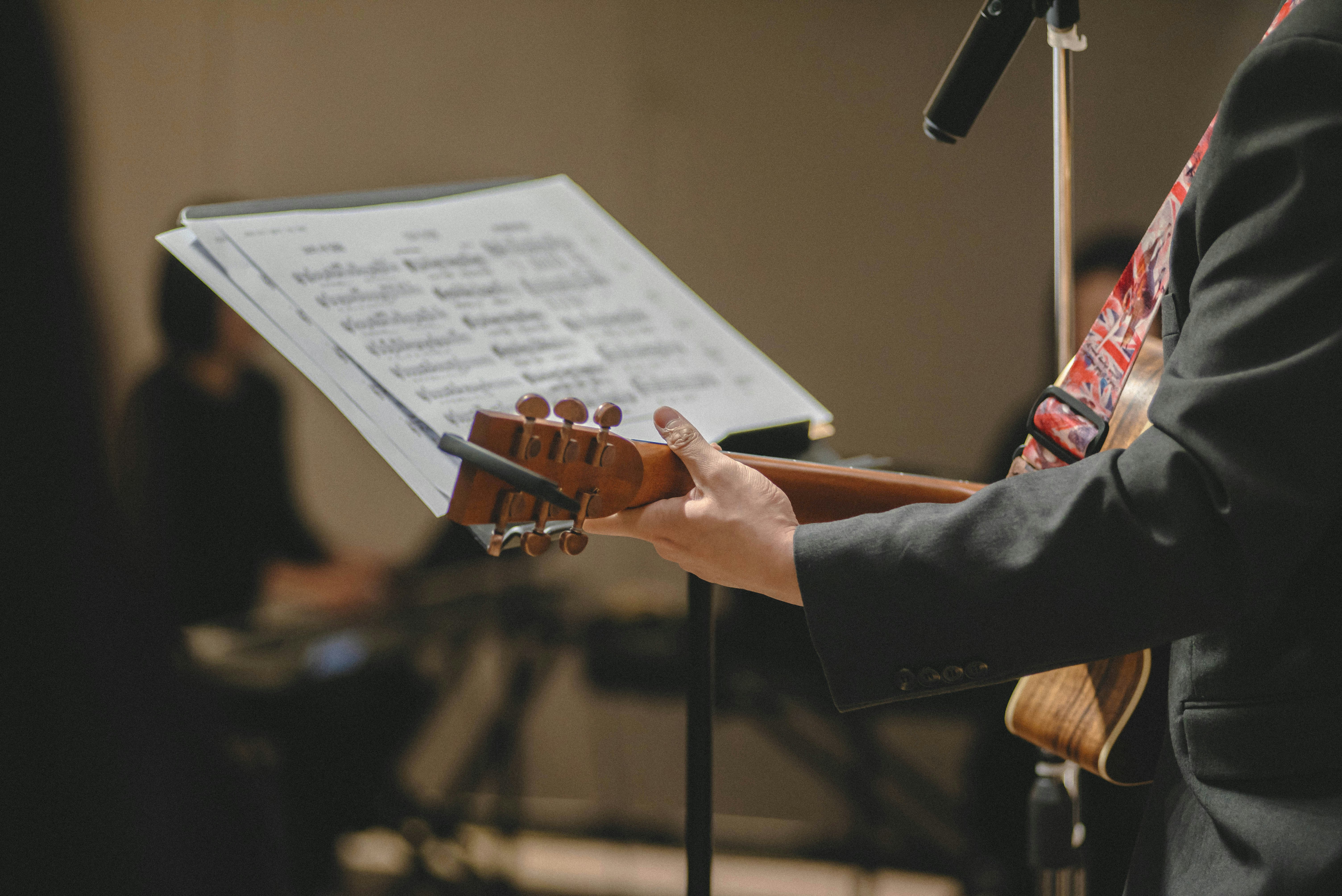 Musician with a guitar stands near a microphone, with a music stand holding sheet music in the foreground.