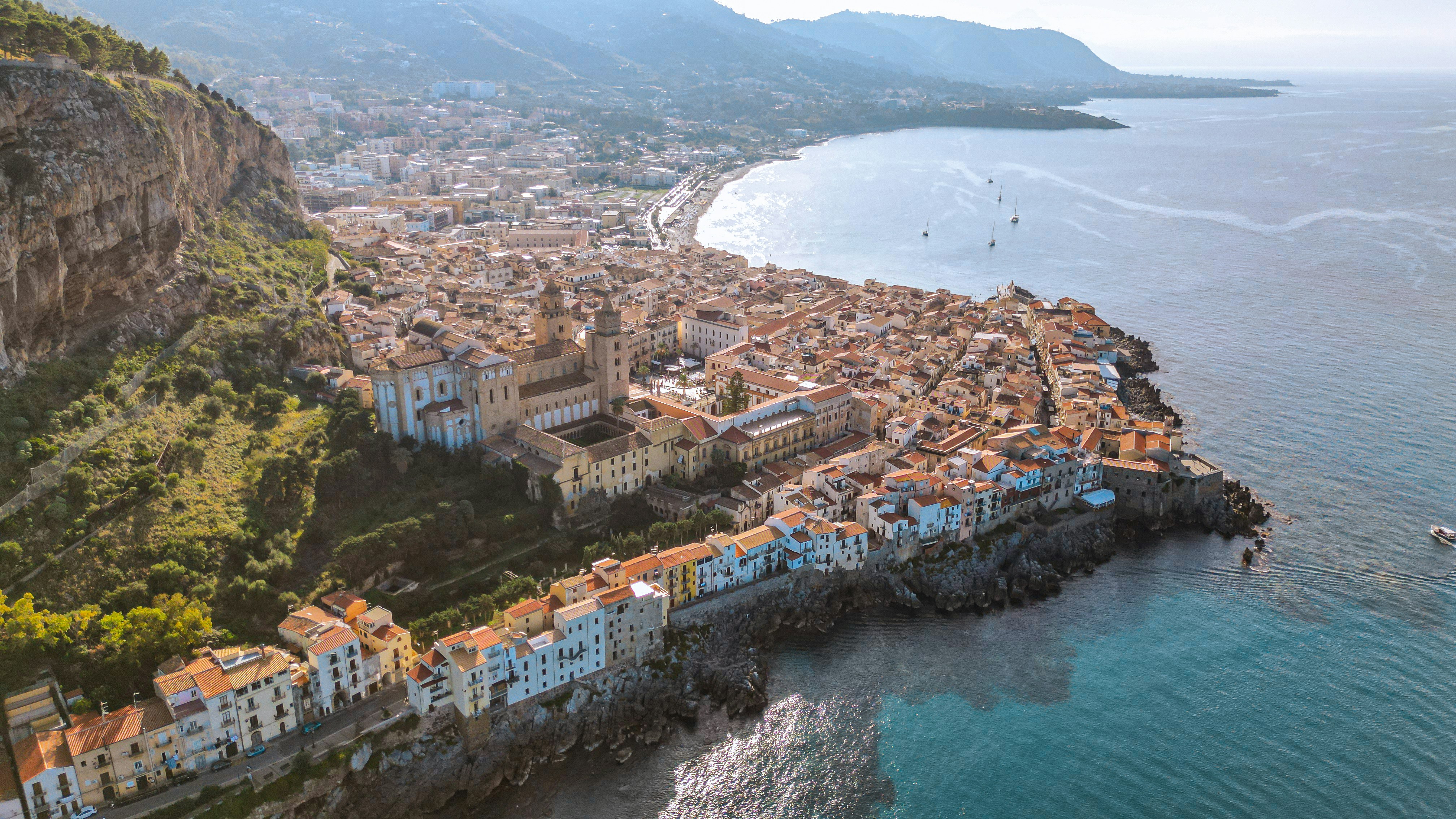 Aerial view of Cefalù's historic buildings nestled between rocky cliffs and the Mediterranean Sea under a clear sky.