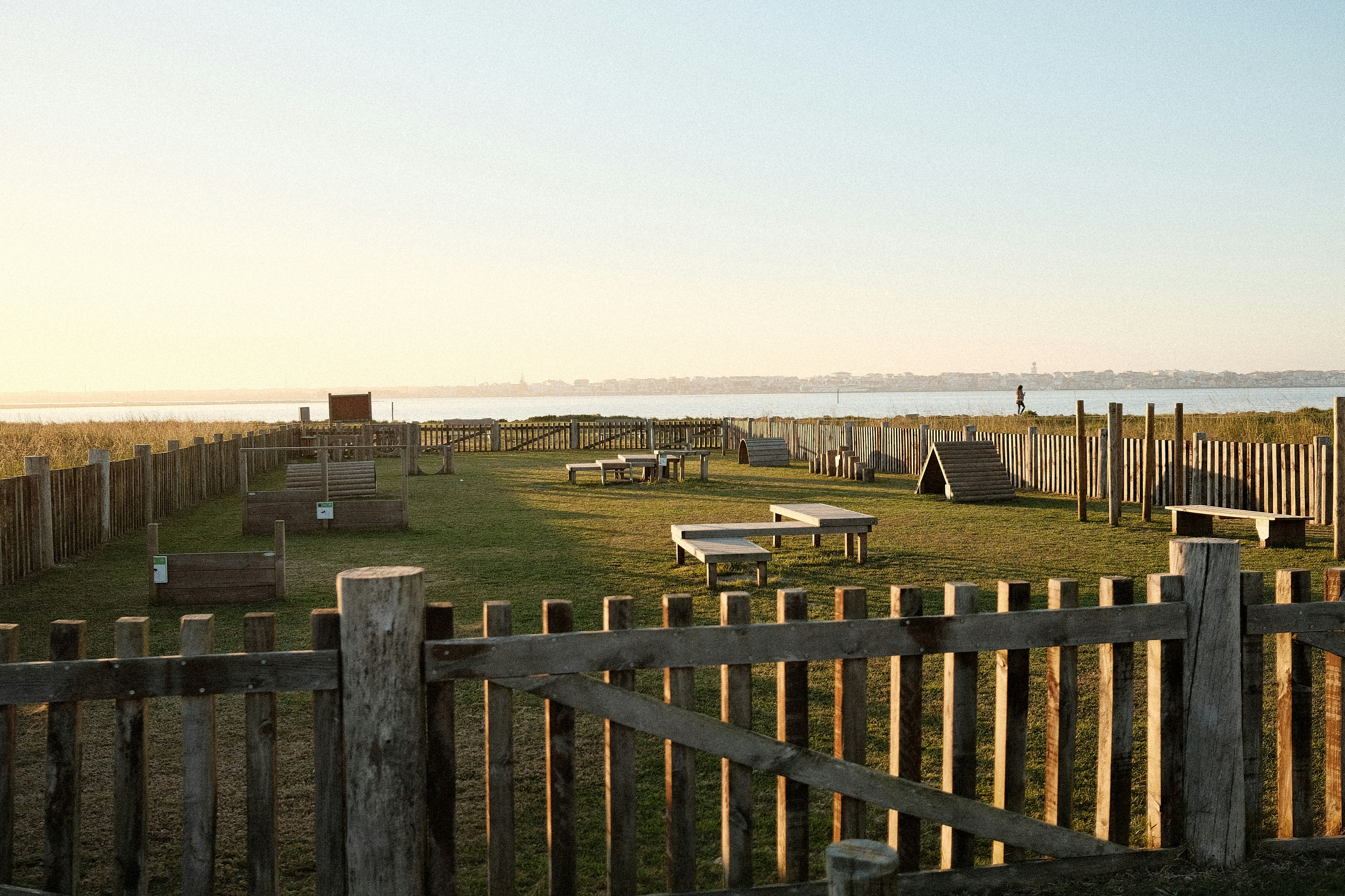 Wooden fences and picnic tables in a grassy area by the sea under a clear sky.