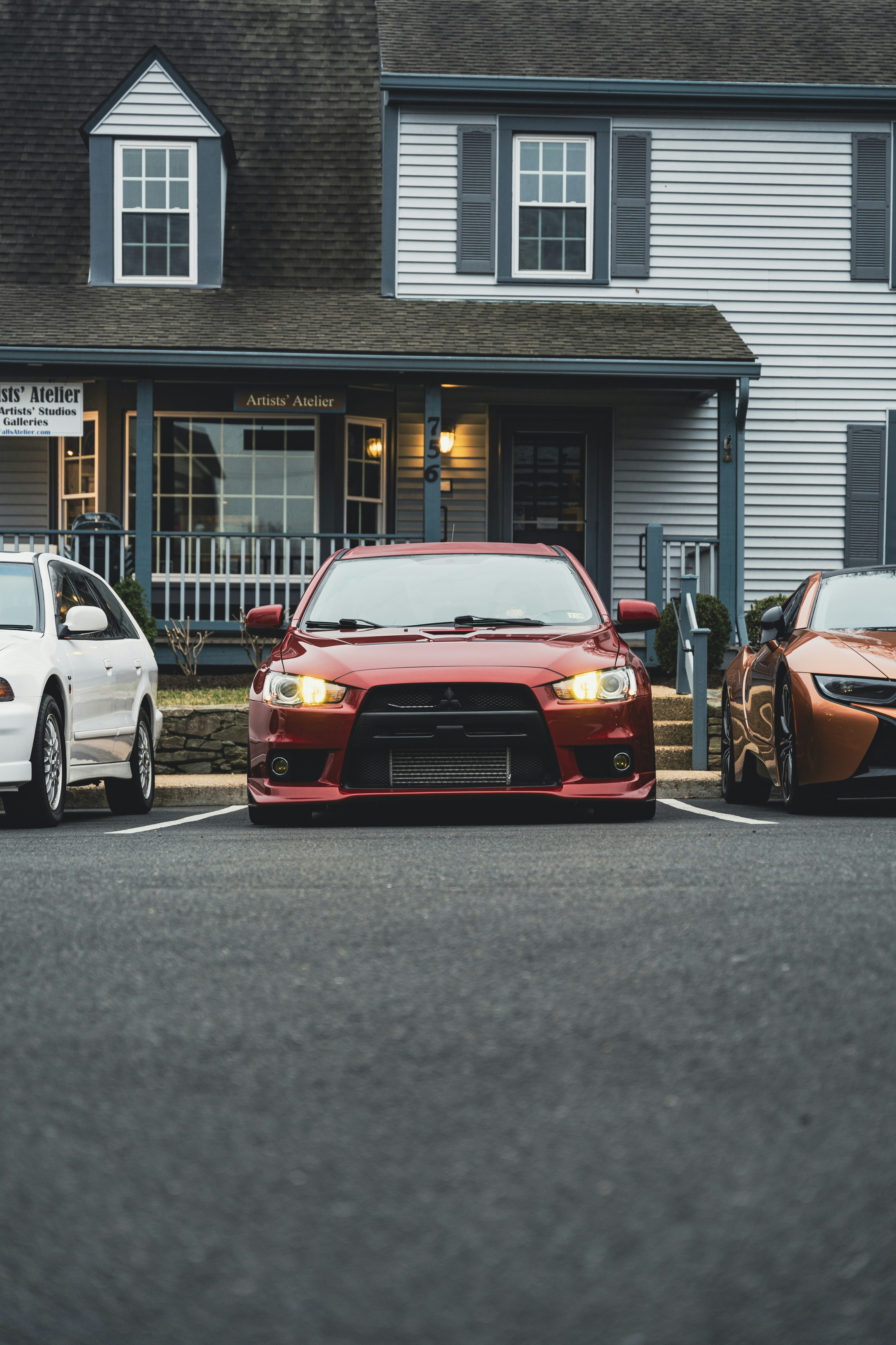 A striking red car is flanked by a white and an orange vehicle in a parking lot, with an artist's studio in the background. The composition highlights the contrast and character of the cars against the charming building.