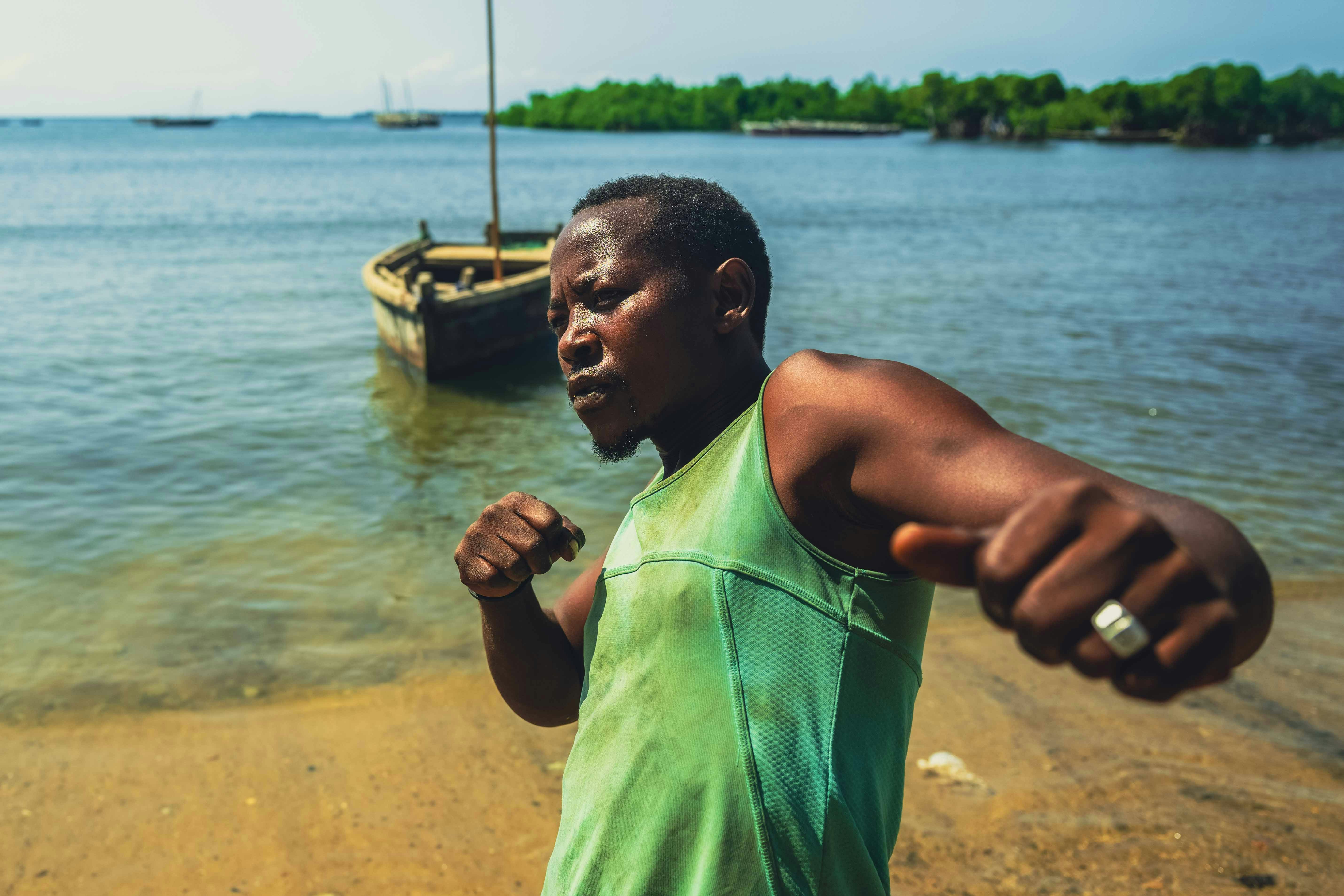 Person in a green shirt throws a punch on a sandy beach with a wooden boat and lush greenery in the background.