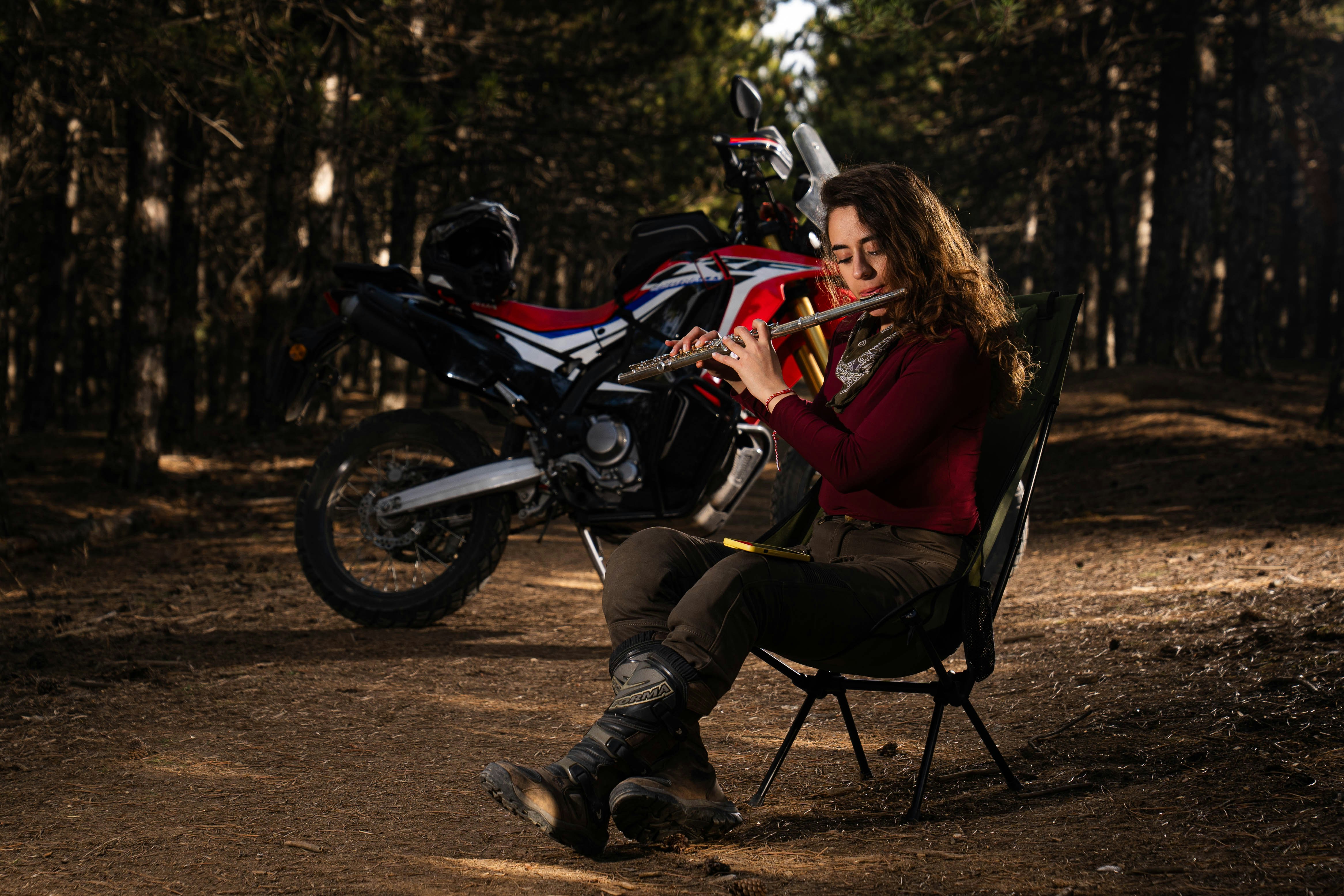 Person playing a flute while seated on a forest path with a motorbike in the background.