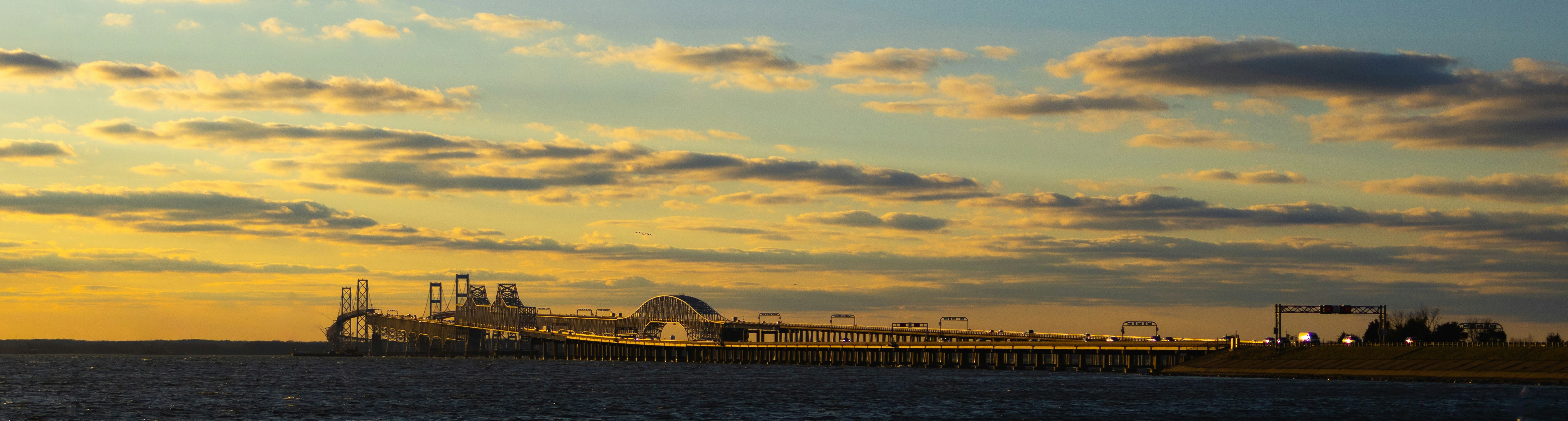 Silhouetted construction cranes against a golden sunset sky with scattered clouds over a calm sea.