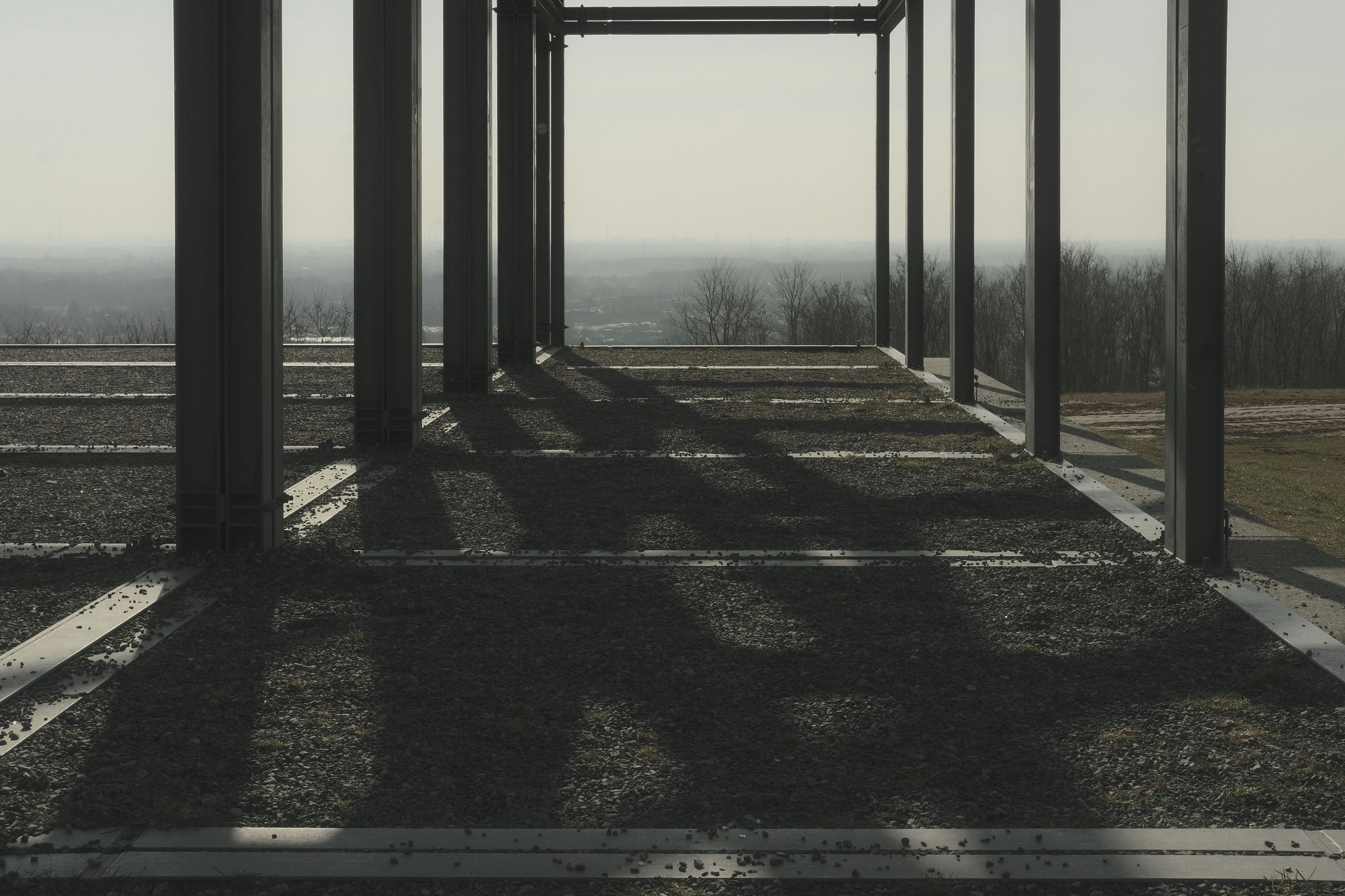 A minimalist architectural framework casting long shadows across a gravel surface, emphasizing the interplay of light and geometry.