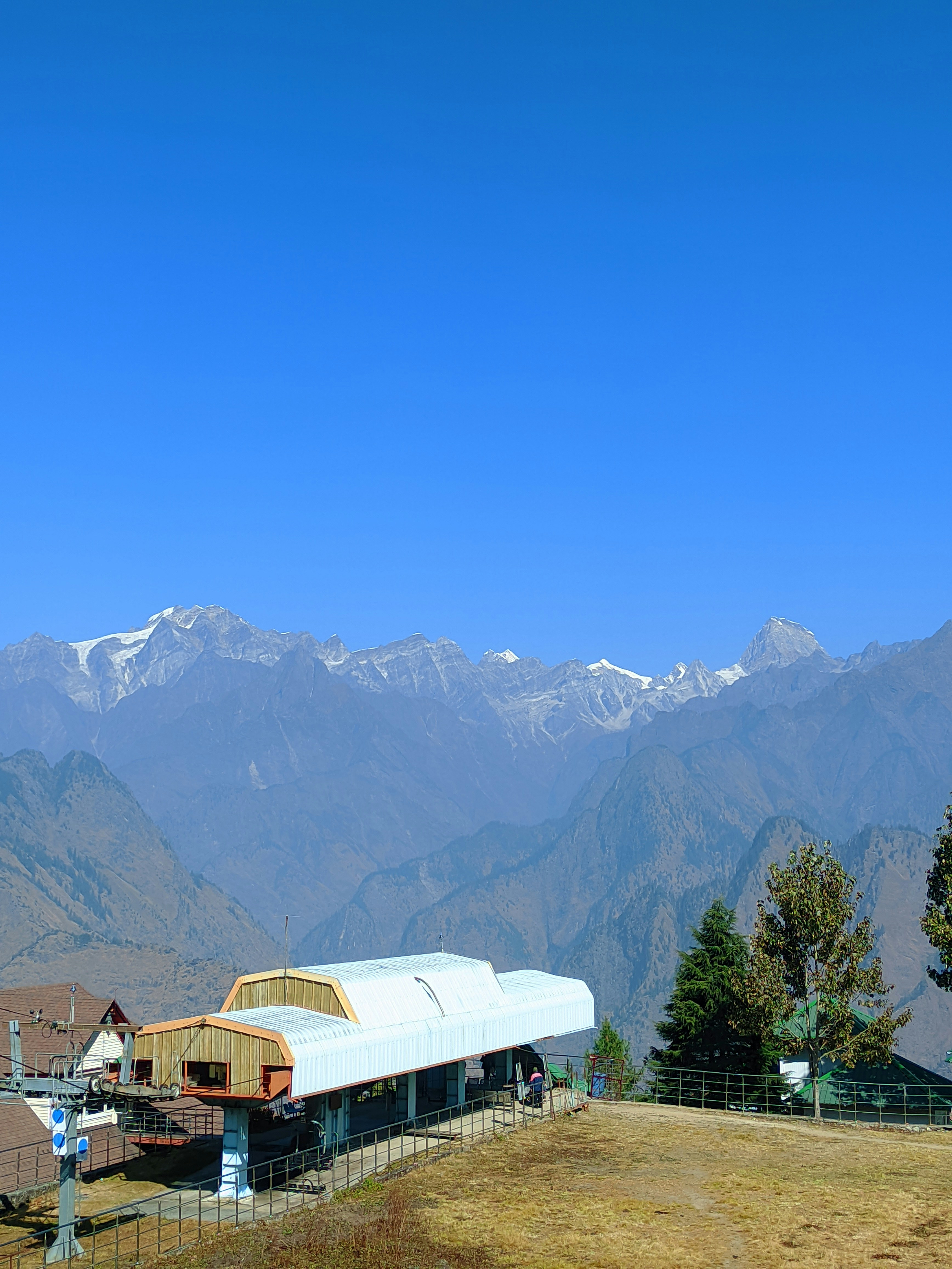 Alpine hilltop lodge with a curved white roof sits on a railing-lined platform, as snow-capped peaks rise behind under a clear blue sky.