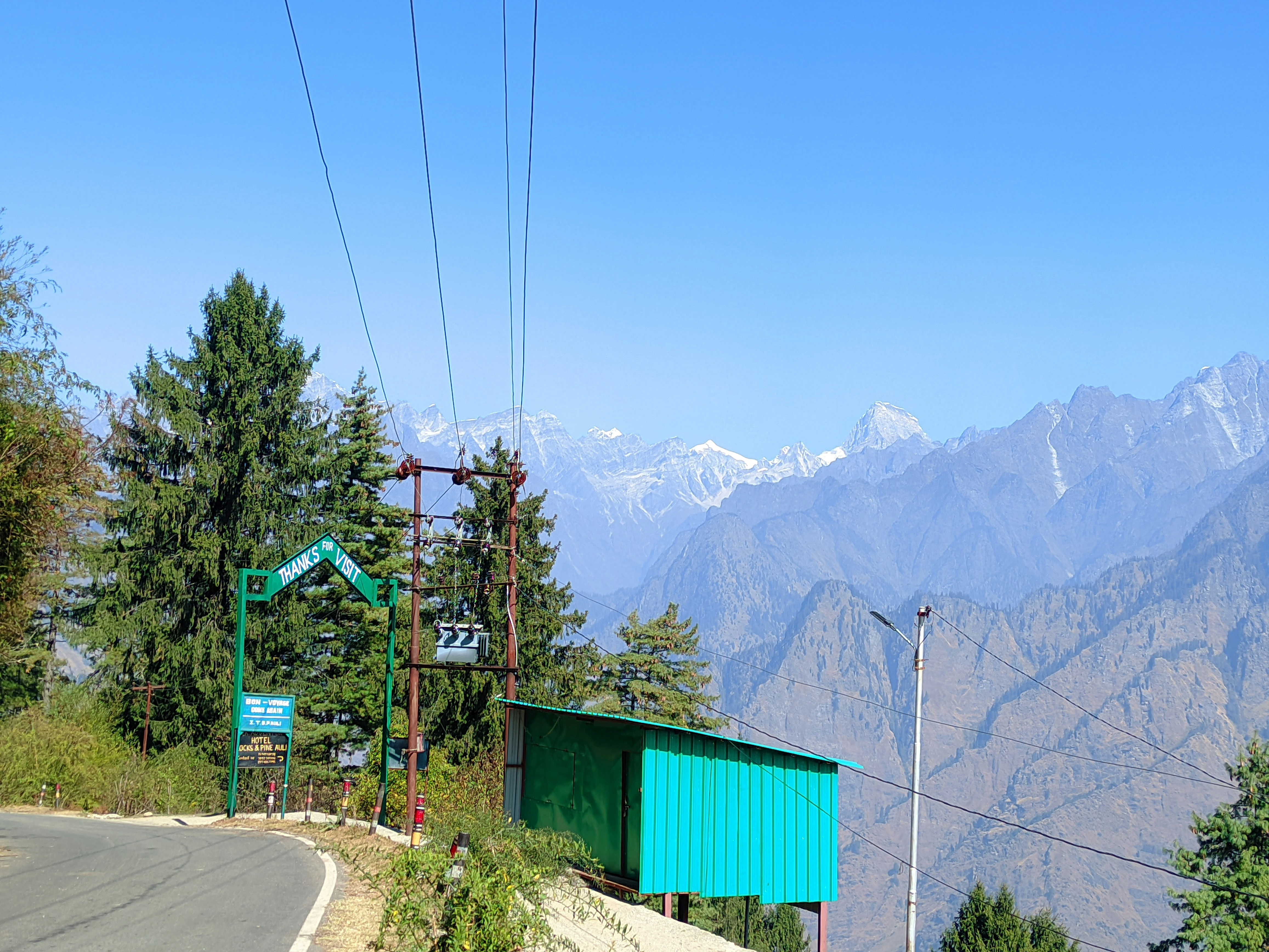 A winding mountain road with a bright turquoise shed and utility poles under a clear blue sky; distant snow-capped peaks frame the scene.
