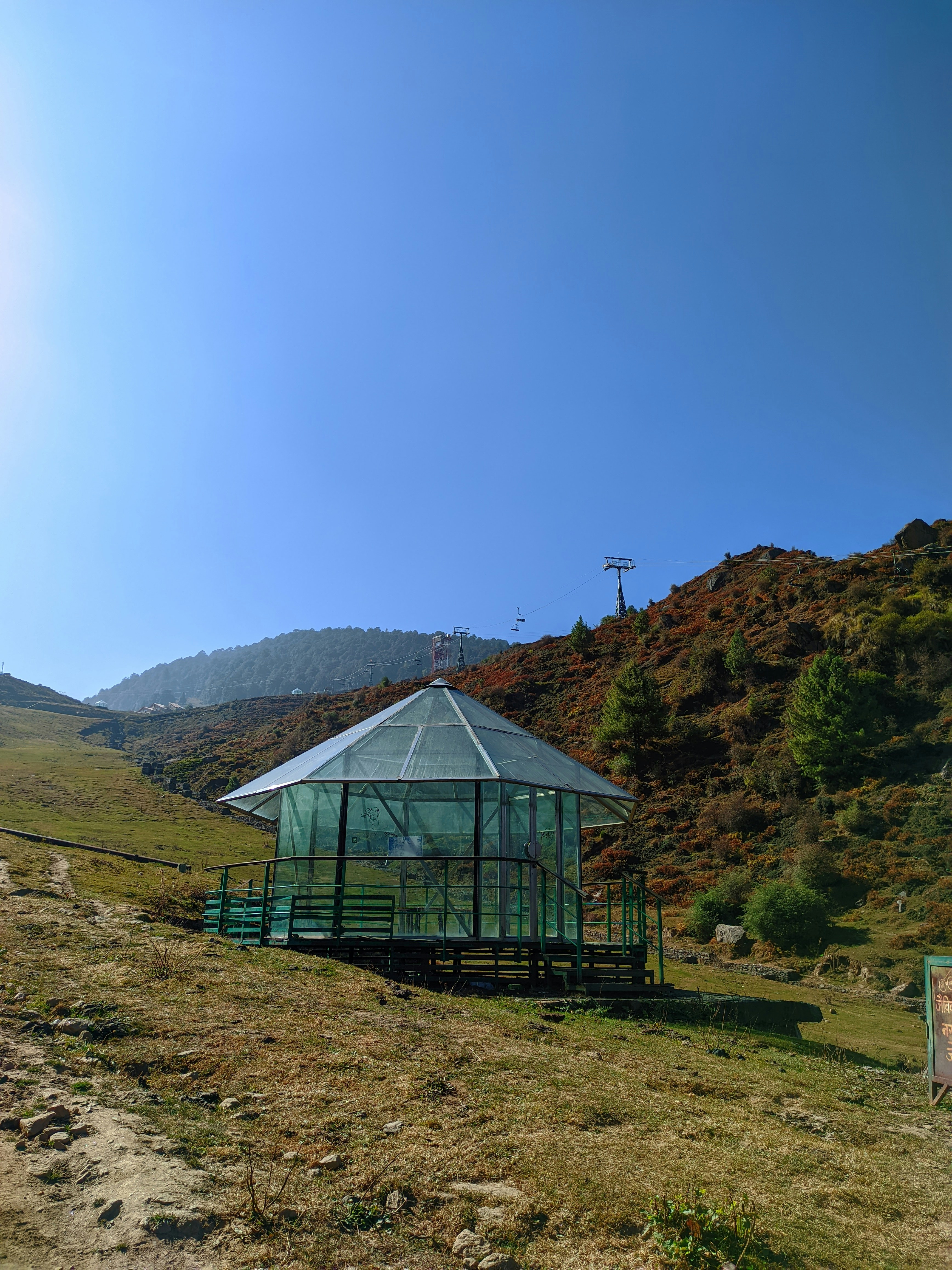 A glass-panel pavilion sits on a grassy hillside with rugged slopes in the background and a clear blue sky above.