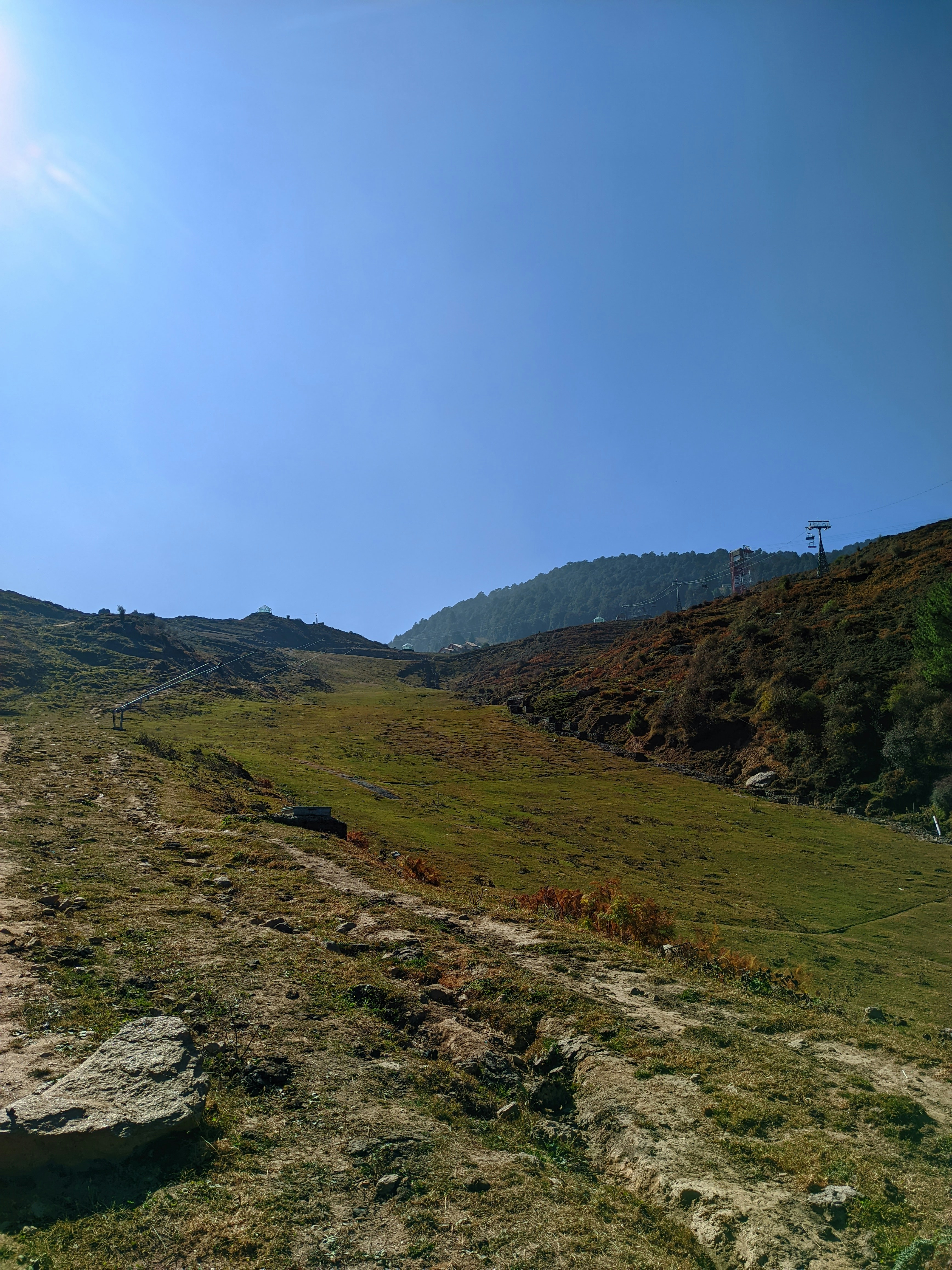 Lush green hillside under a clear blue sky, showcasing a winding path leading up the slope. The sunlight casts a gentle glow on the landscape.