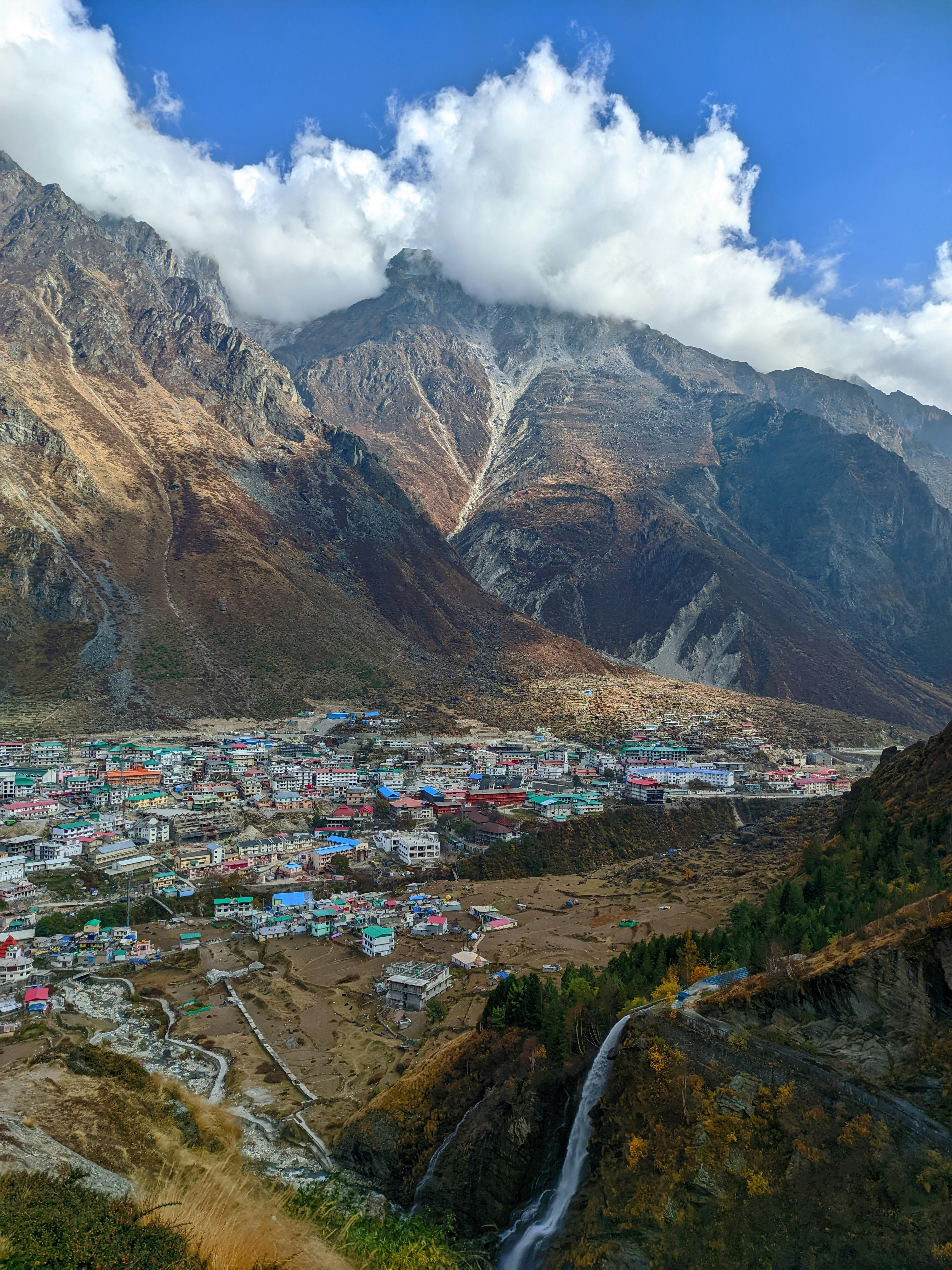 Mountain town nestled beneath towering peaks and drifting clouds, with a waterfall in the foreground.