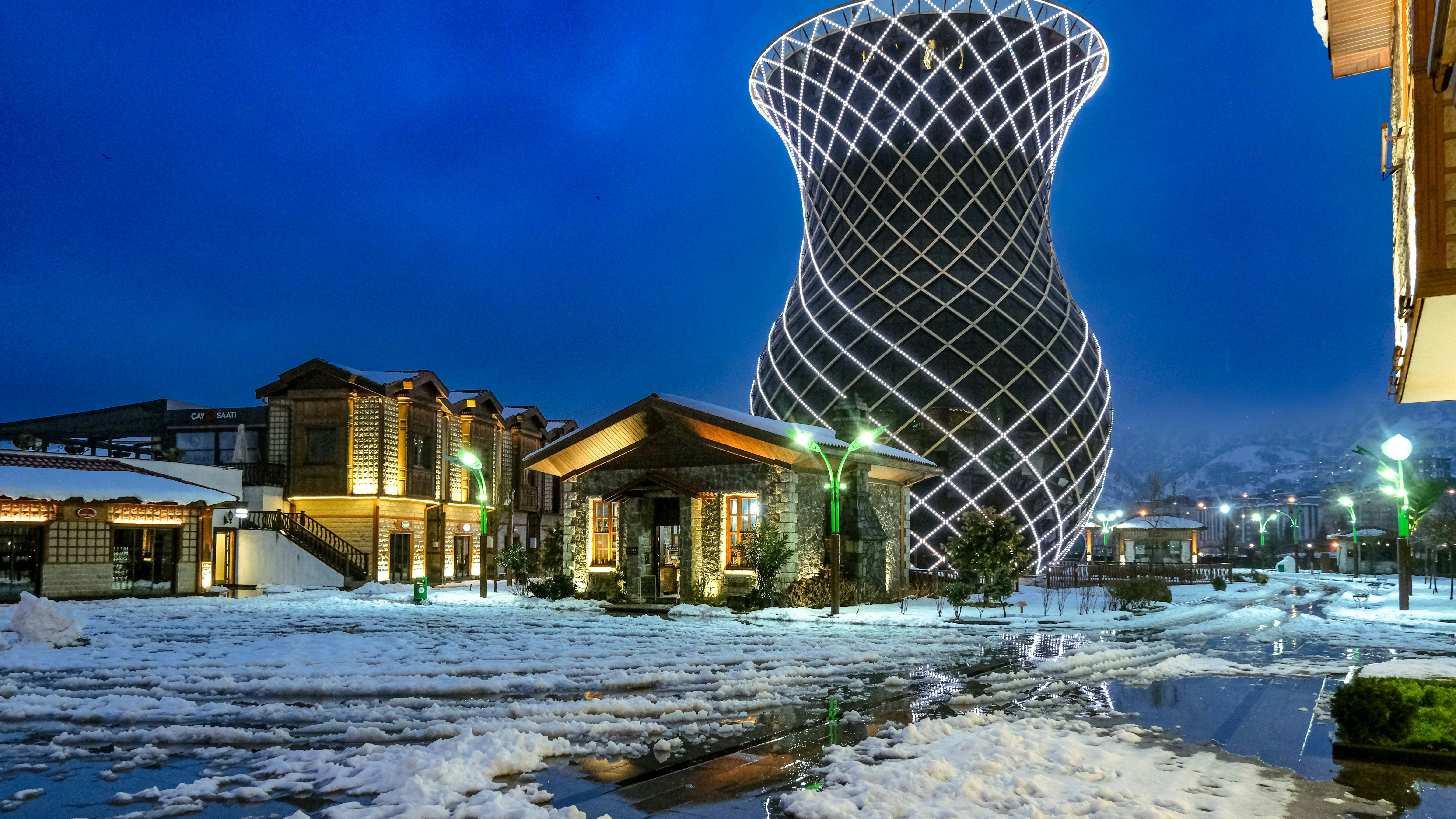 Illuminated lattice tower against a deep blue evening sky surrounded by snow and modern buildings.
