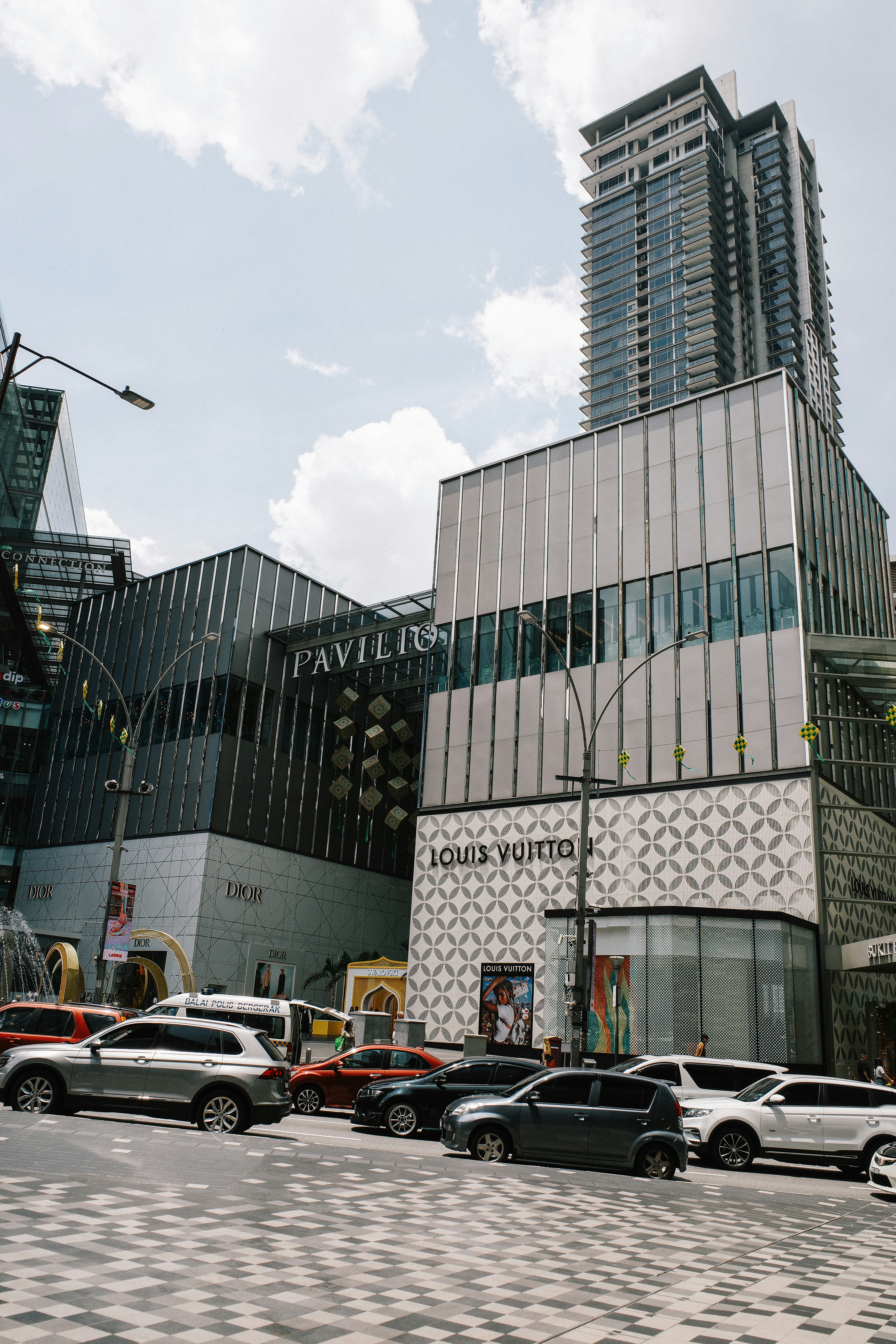 Modern shopping complex and skyscraper against a bright sky, with cars lining the street.