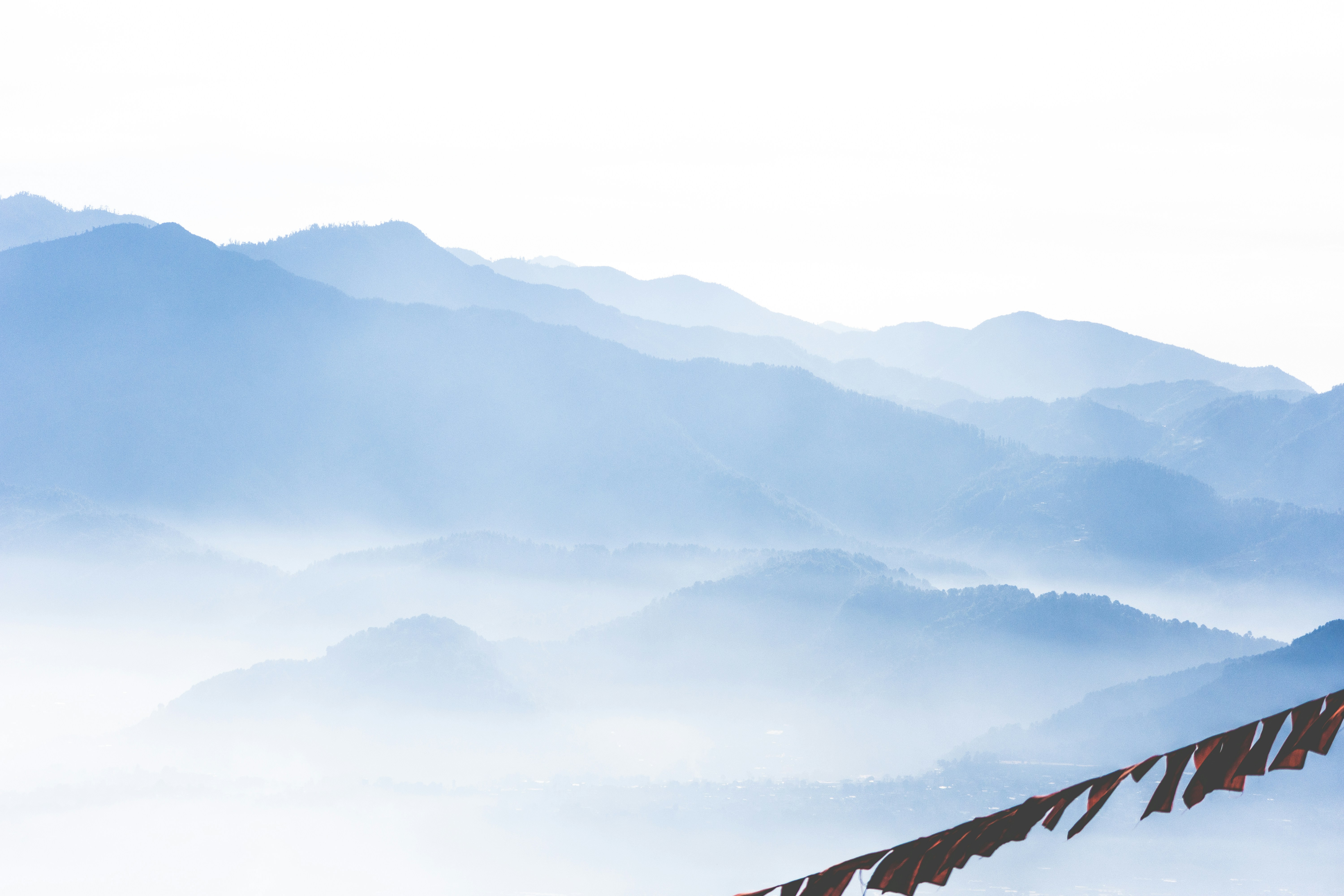 A layered mountain range fades into a misty horizon, with prayer flags fluttering in the foreground. The soft blue hues create a serene and spiritual atmosphere, hinting at a peaceful, remote location.