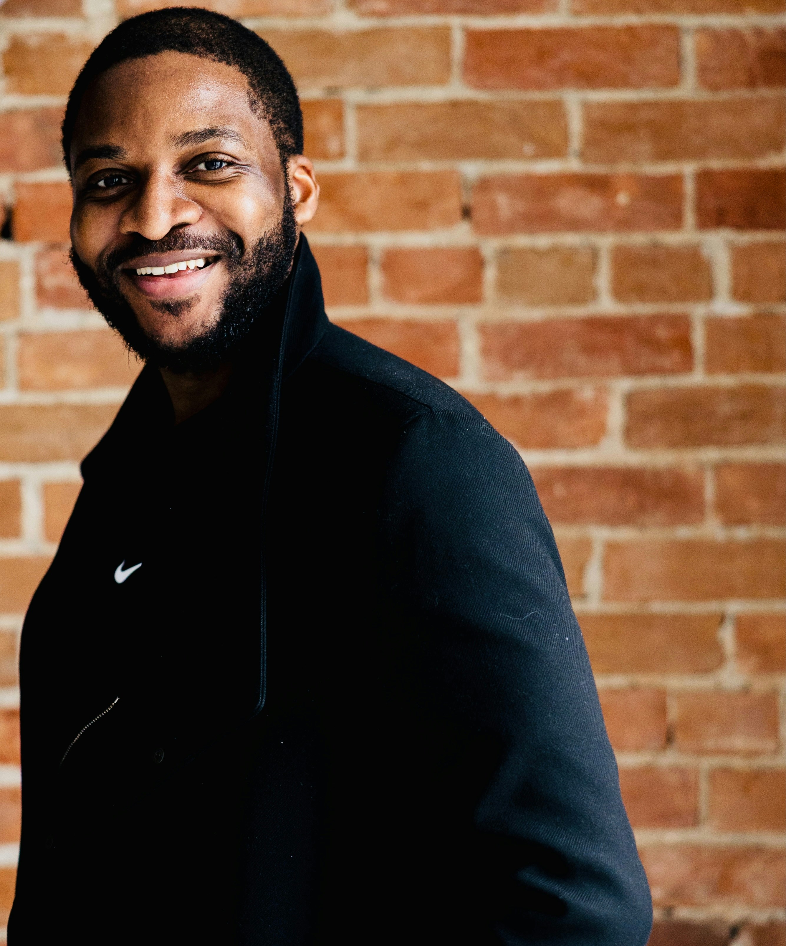 Smiling man in a dark coat standing in front of a textured brick wall.