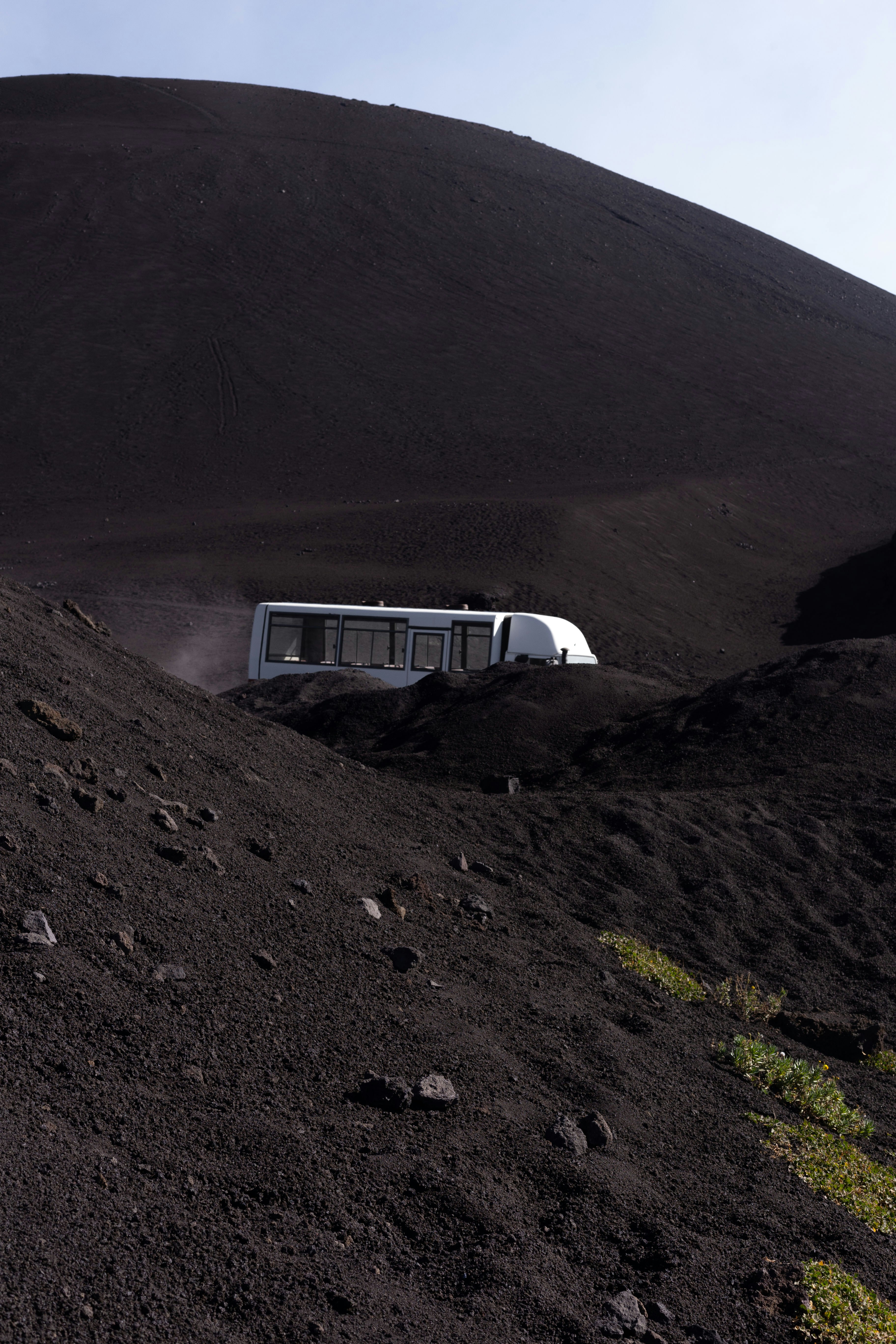 White vehicle traversing a rugged, dark volcanic landscape with sparse vegetation and a clear sky.
