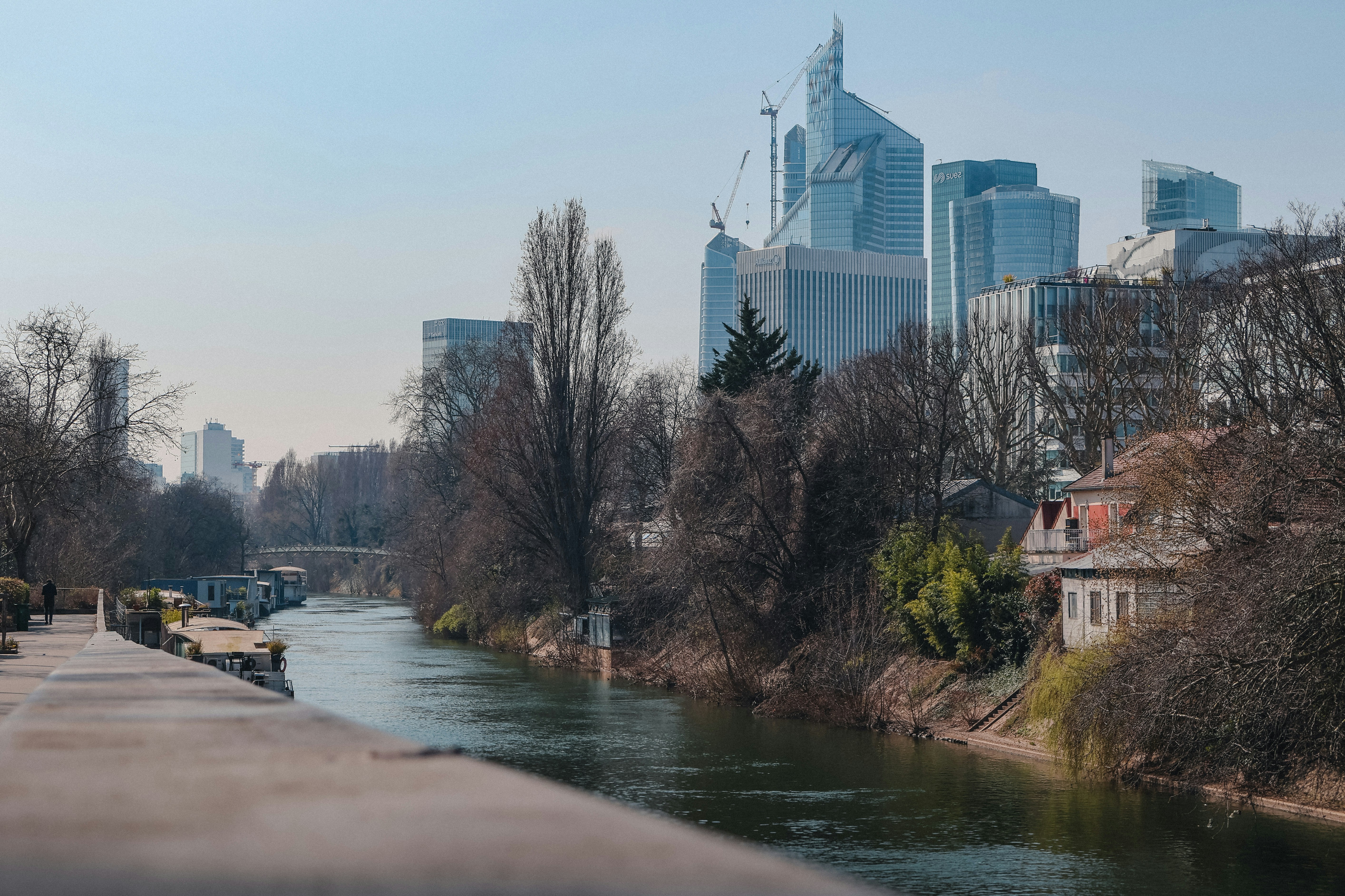 Modern skyscrapers rise above a tranquil river bordered by trees and quaint houses.