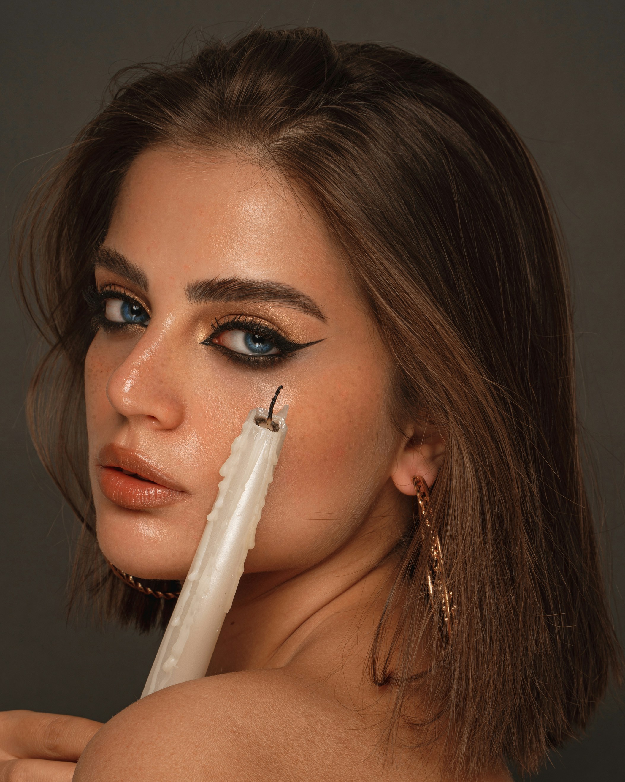 A woman with striking blue eyes and bold eyeliner holds a white candle close to her face, set against a dark background.