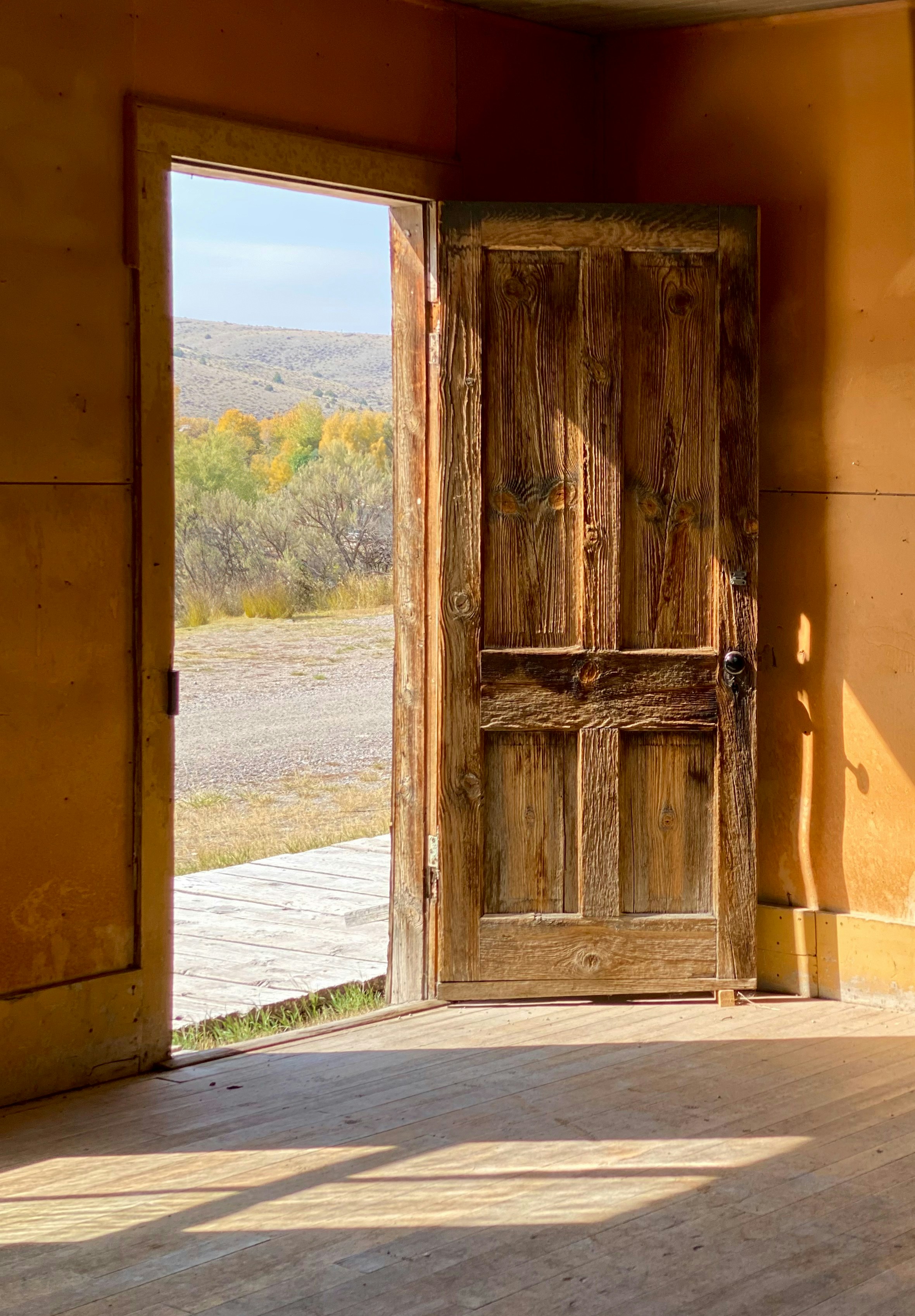 Weathered wooden door ajar, revealing a glimpse of vibrant autumn foliage outside. The warm tones of the interior contrast with the natural landscape beyond.