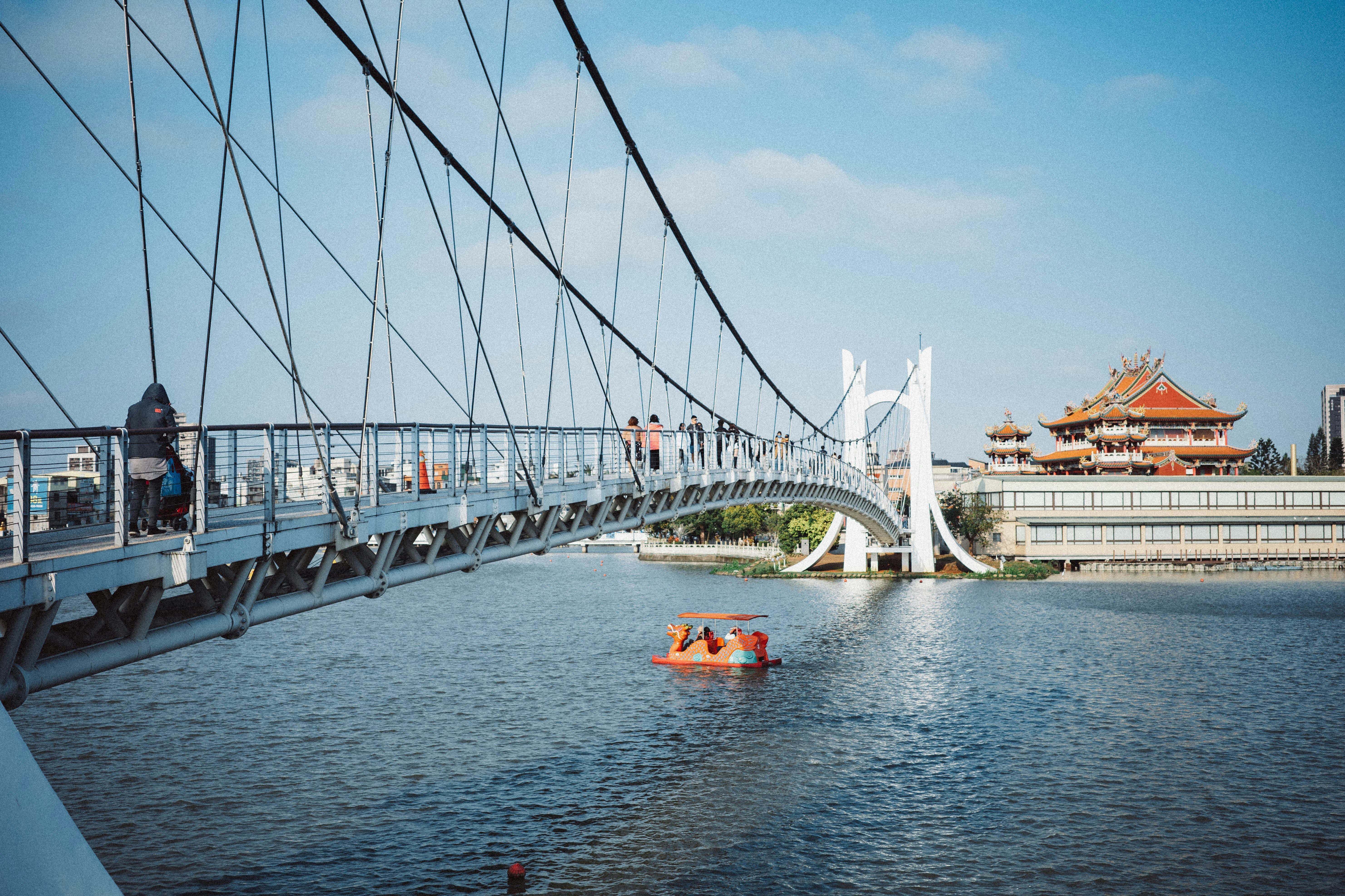 Suspension bridge arches over a river with a traditional building in the background under a clear blue sky.