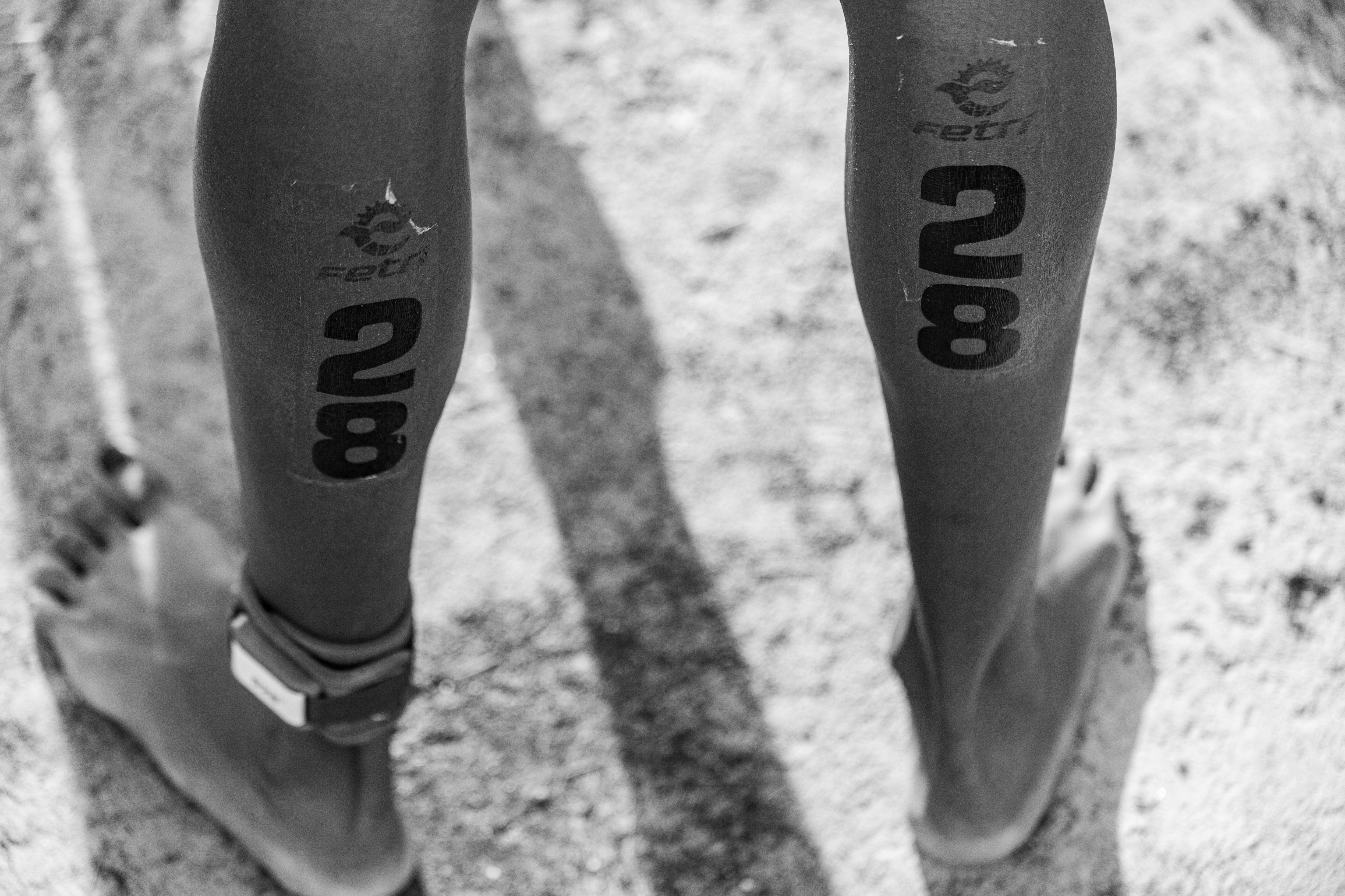Athlete's legs adorned with race numbers and timing chip, standing on sandy terrain. The monochrome tones emphasize the intensity of competition.