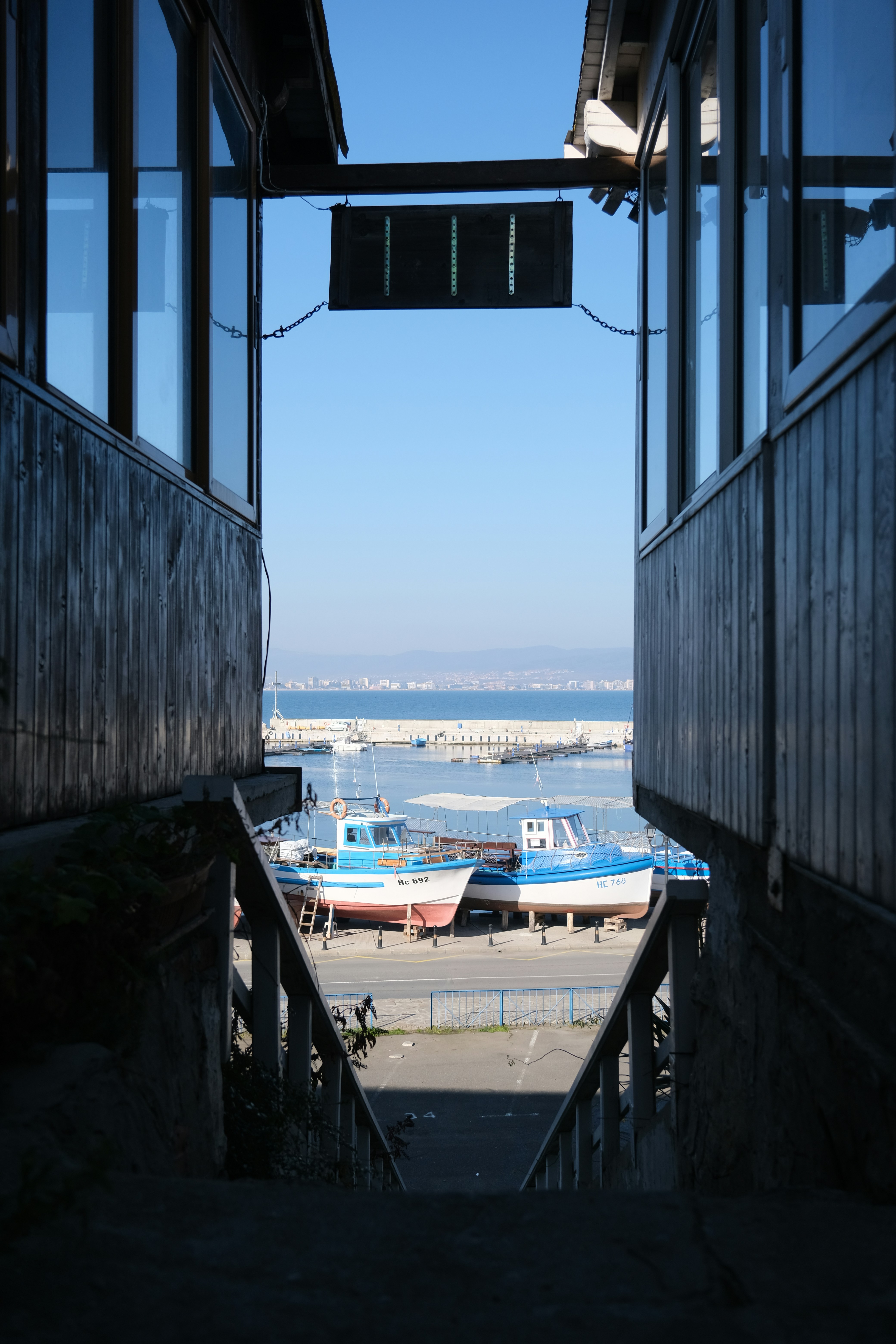 View of fishing boats docked in a harbor, framed by the wooden architecture of nearby buildings. Clear blue sky enhances the tranquil atmosphere.