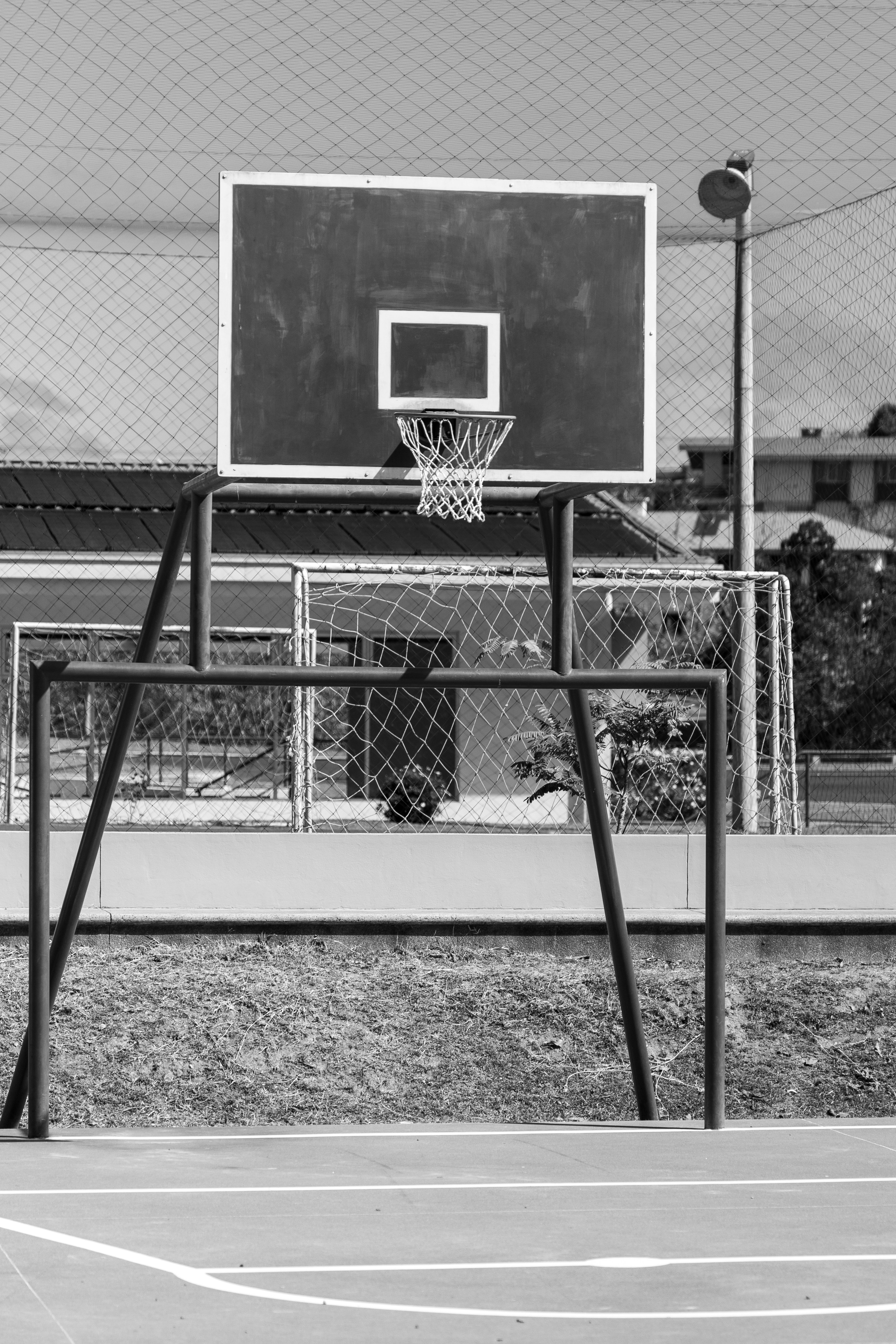 A black-and-white photograph capturing a basketball hoop in an outdoor court, evoking a sense of nostalgia and simplicity. The weathered backboard and net contrast with the surrounding urban environment, enclosed by fencing. The image's monochrome tones highlight the geometric lines and textures, emphasizing the raw essence of street basketball. A solitary yet inviting scene, it reflects the universal appeal of the sport and the quiet moments in an empty court.