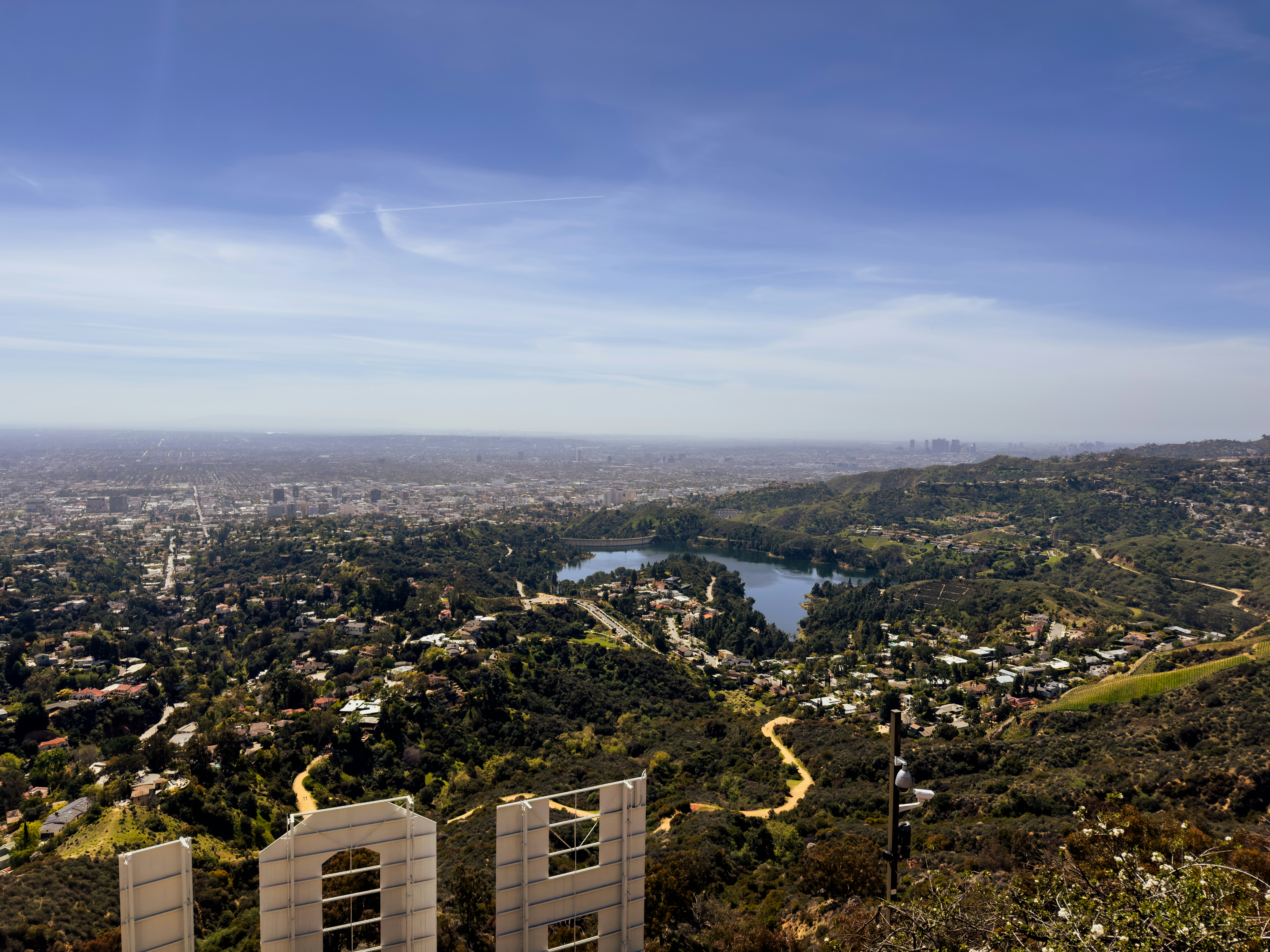 Expansive view of a cityscape with a serene lake nestled among green hills, framed by iconic Hollywood sign letters in the foreground.