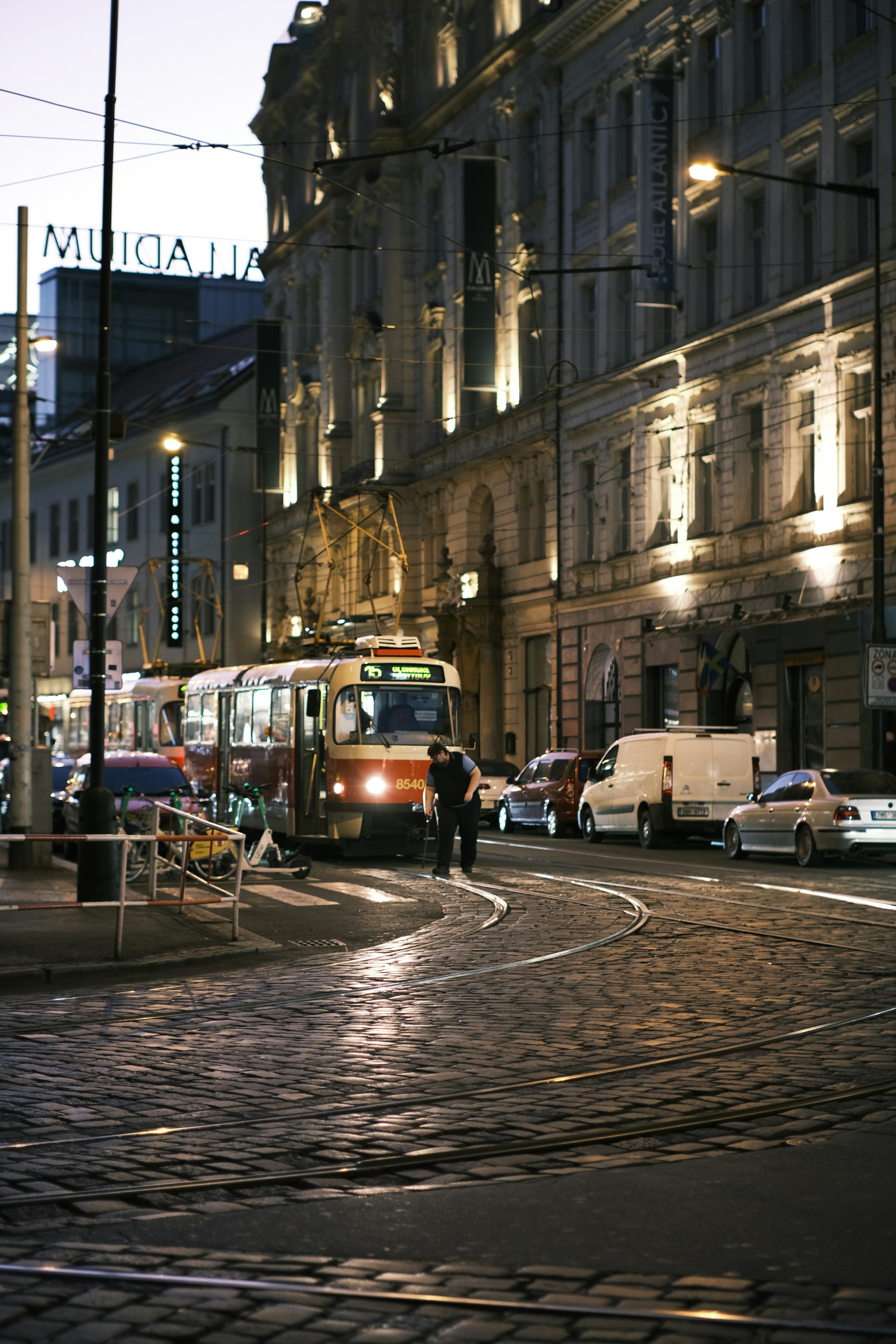 Evening urban life captured on a busy street illuminated by city lights and a tram waiting for operator to switch lanes with a crowbar. | A tram car is moving on a cobblestone street.