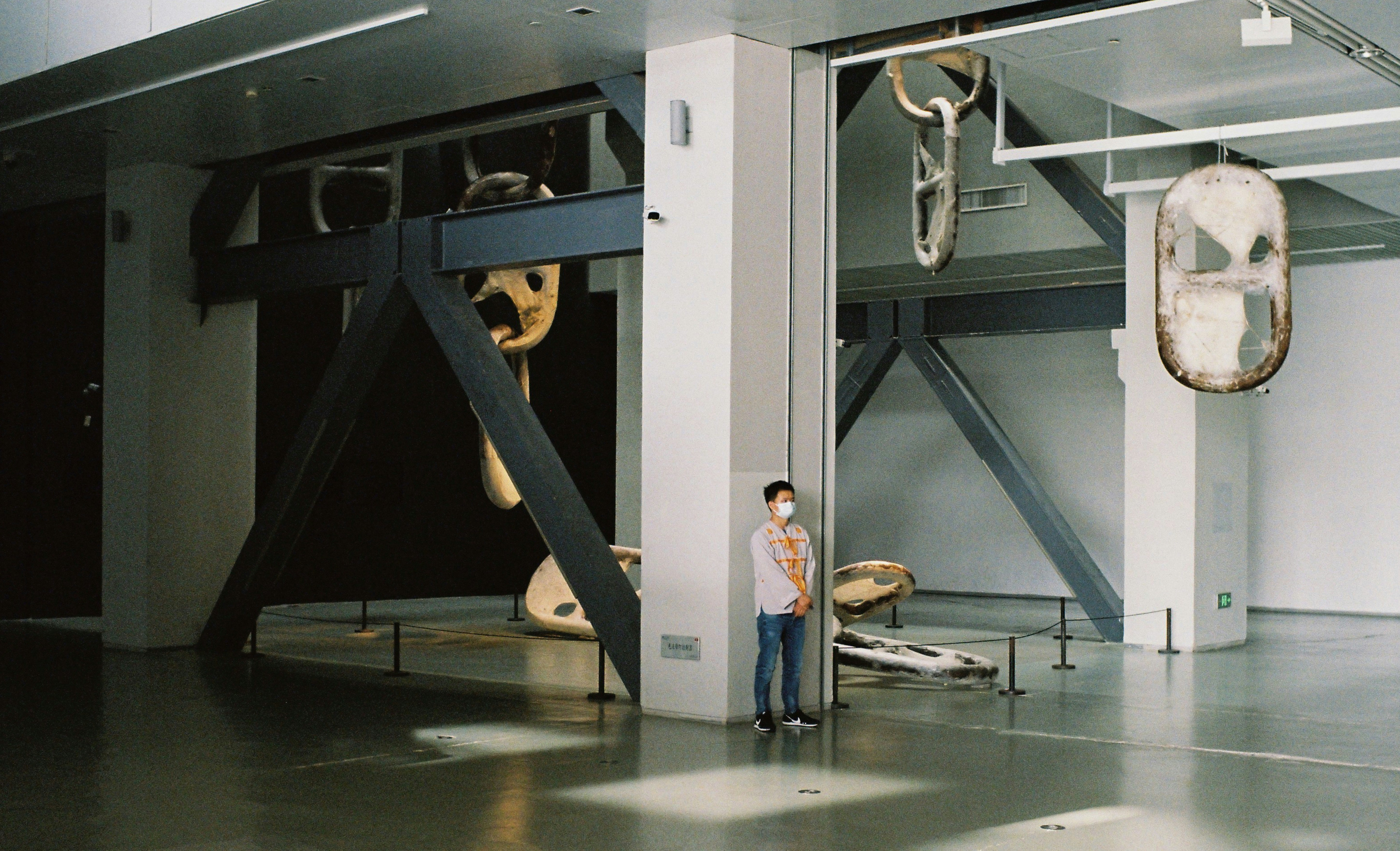 Solitary visitor in a stark industrial gallery stands near a white pillar, surrounded by oversized mechanical sculptures and hanging forms.