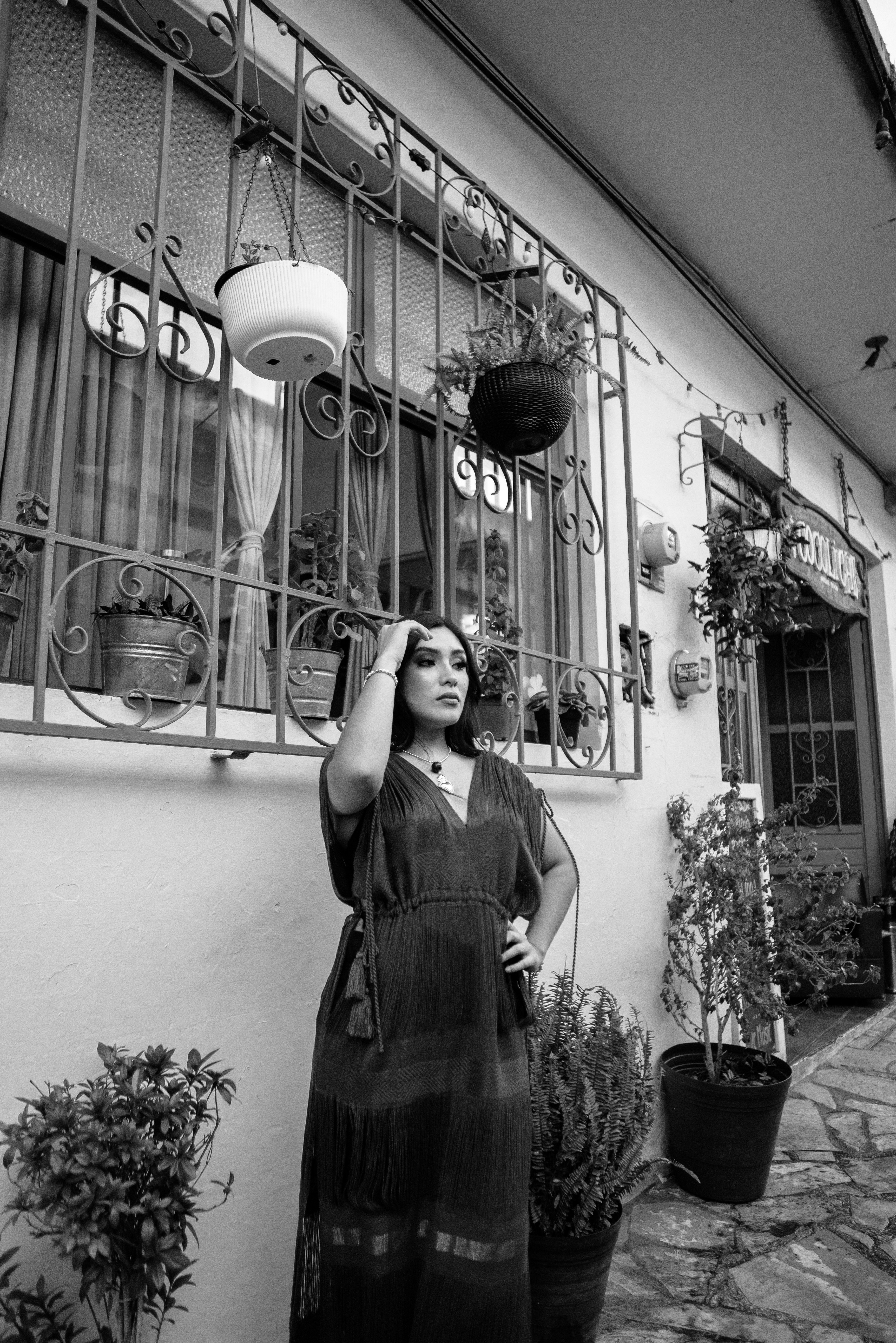 Woman in a long dress stands thoughtfully by a window adorned with hanging plants on a city street.