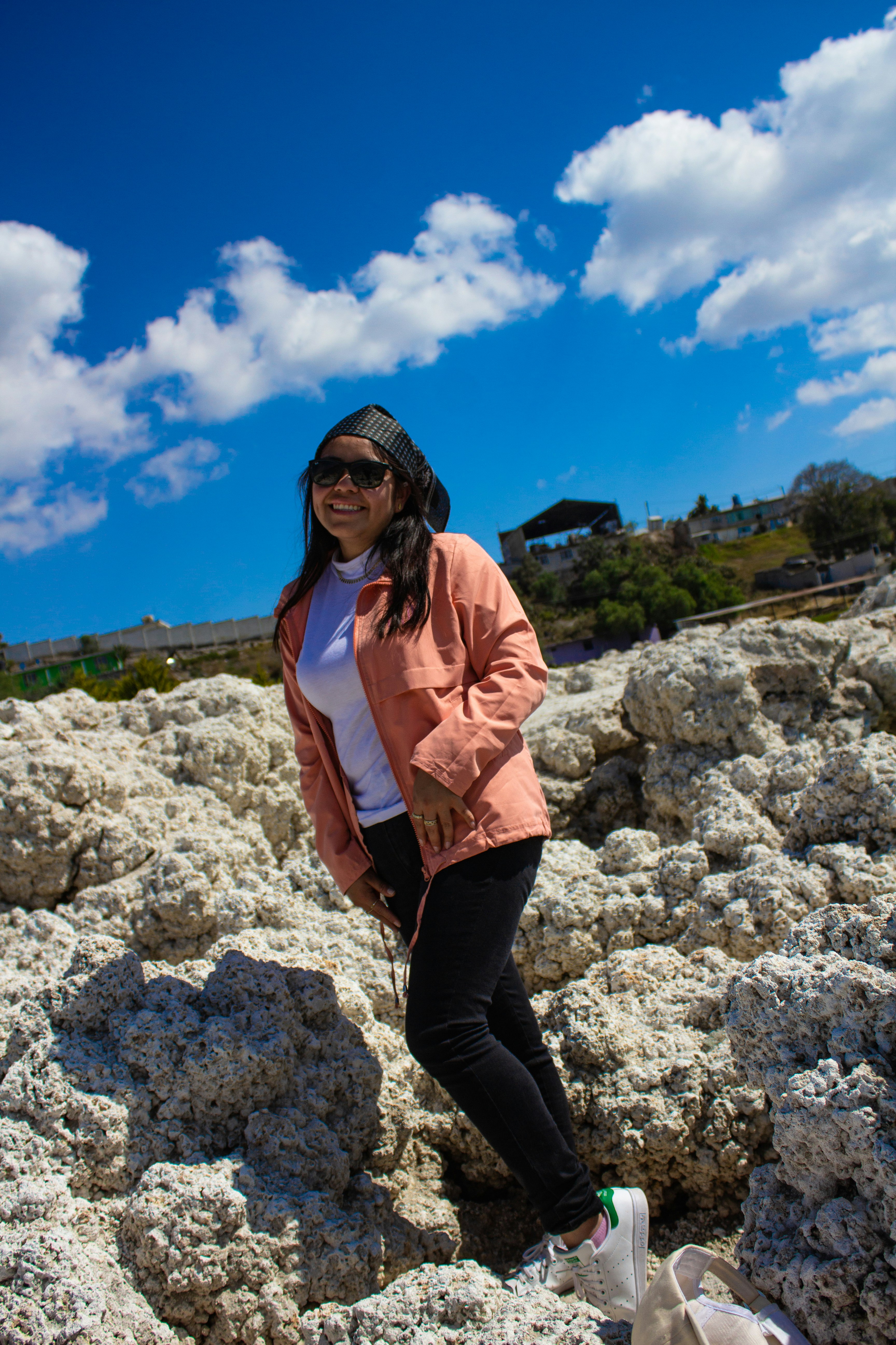Smiling individual in a coral jacket standing on rugged white rocks under a bright blue sky with scattered clouds.