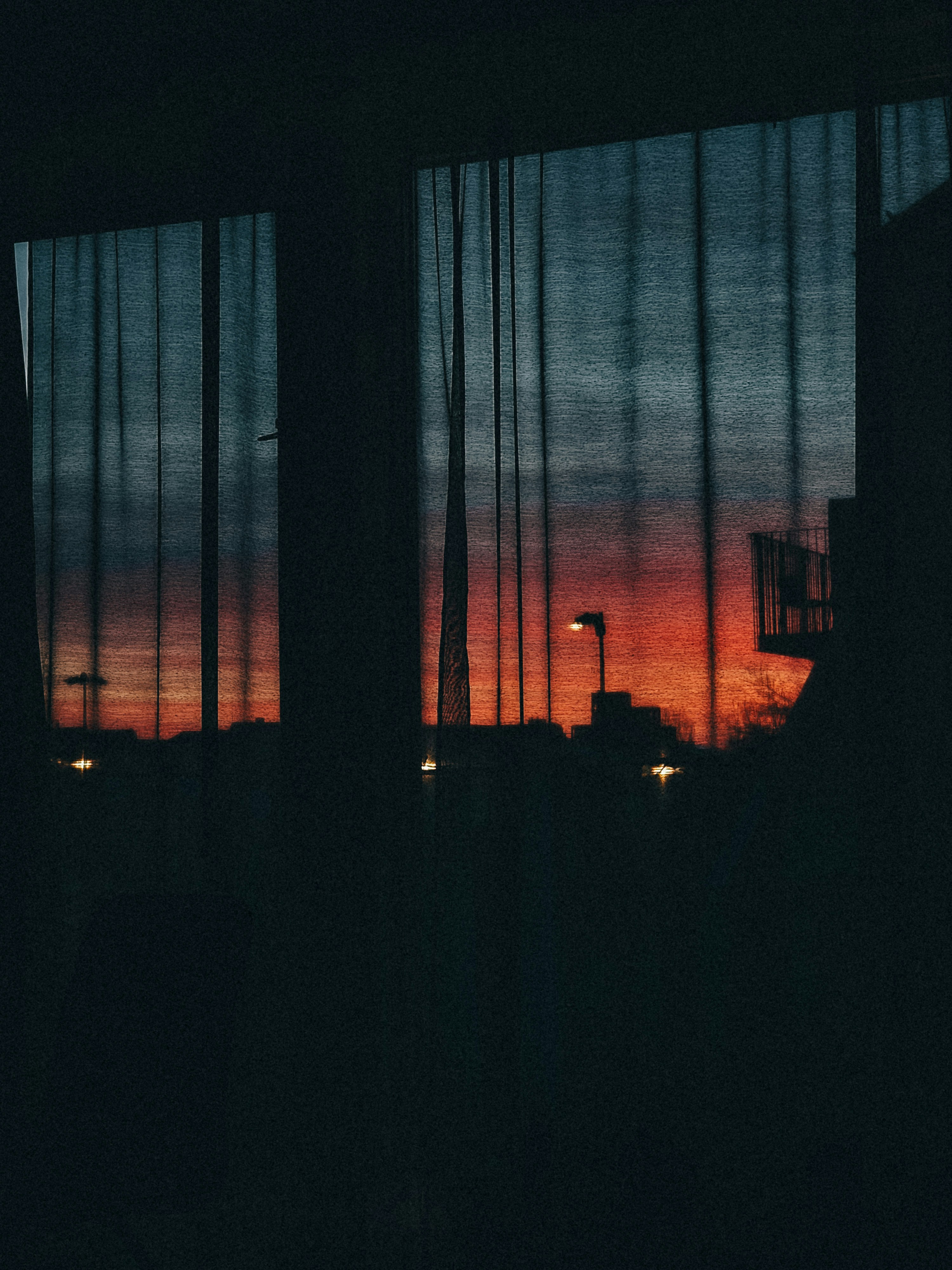 Dark interior frames a sunset-lit cityscape through vertical blinds. A warm orange horizon and silhouetted streetlights outline the skyline.