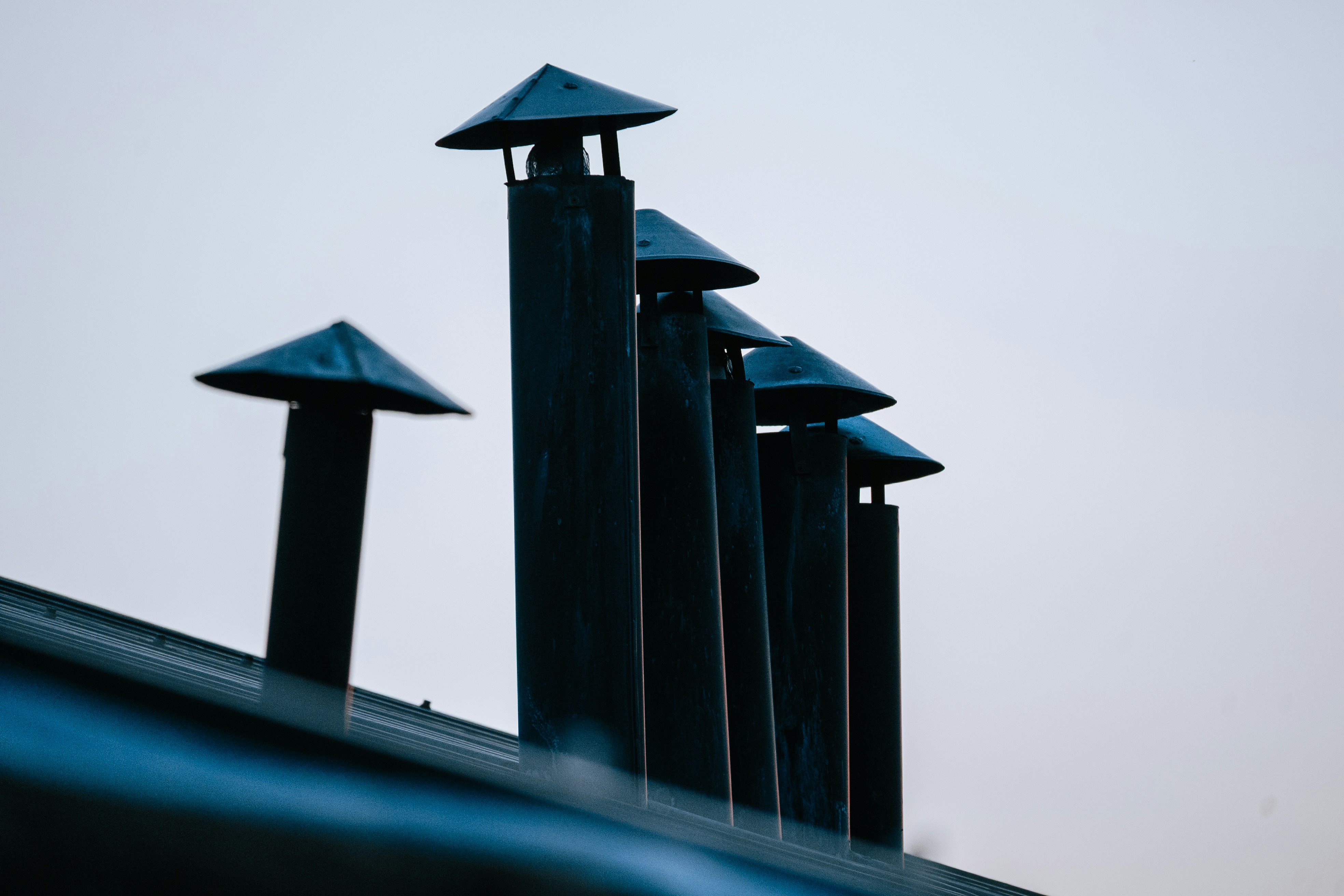 Row of industrial metal chimneys with conical caps against a pale sky, creating a geometric silhouette. | Black chimneys on a roof against the sky.