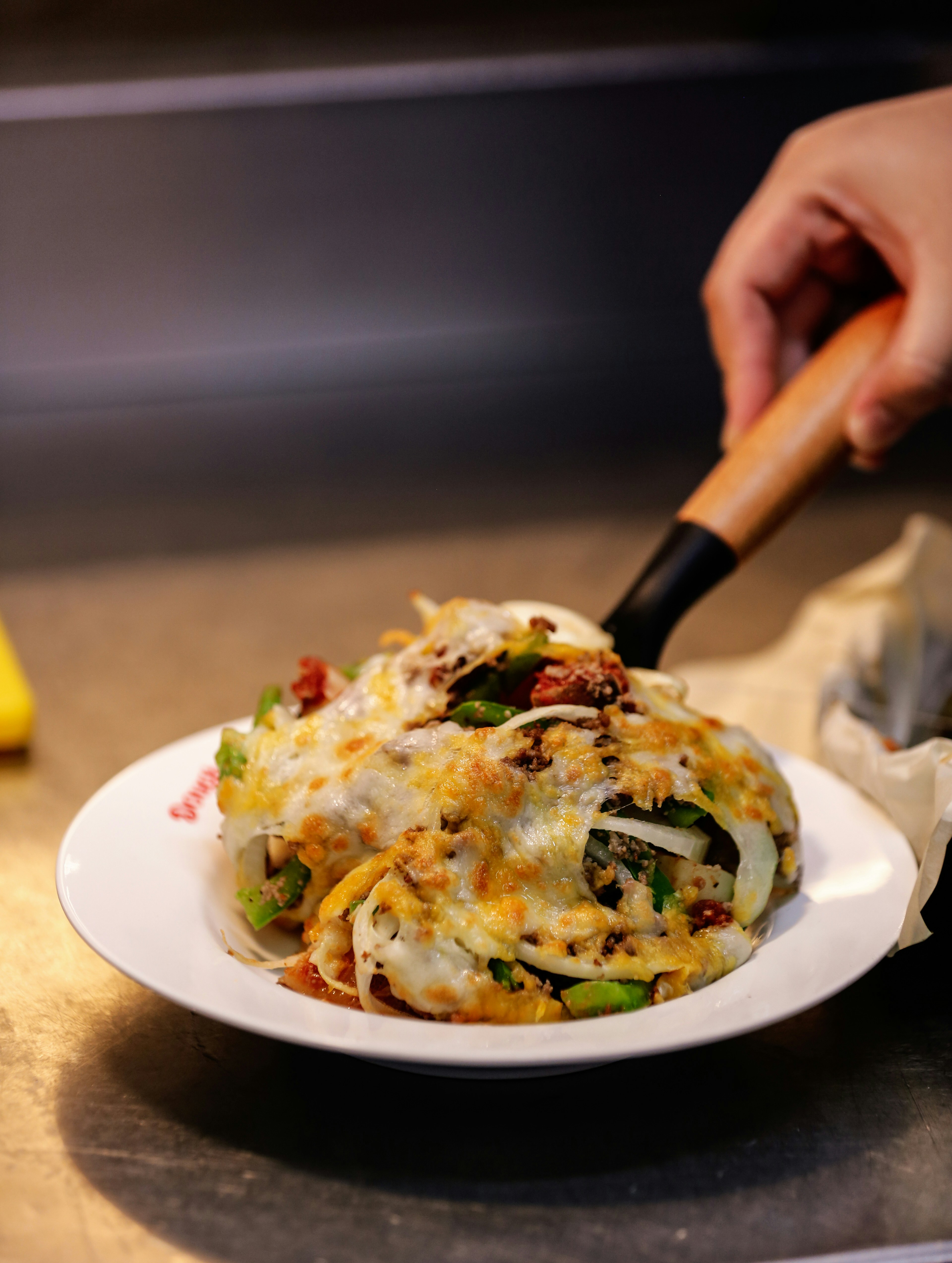 A hand serving a deliciously cheesy baked dish topped with colorful vegetables on a white plate, showcasing the artistry of home cooking.