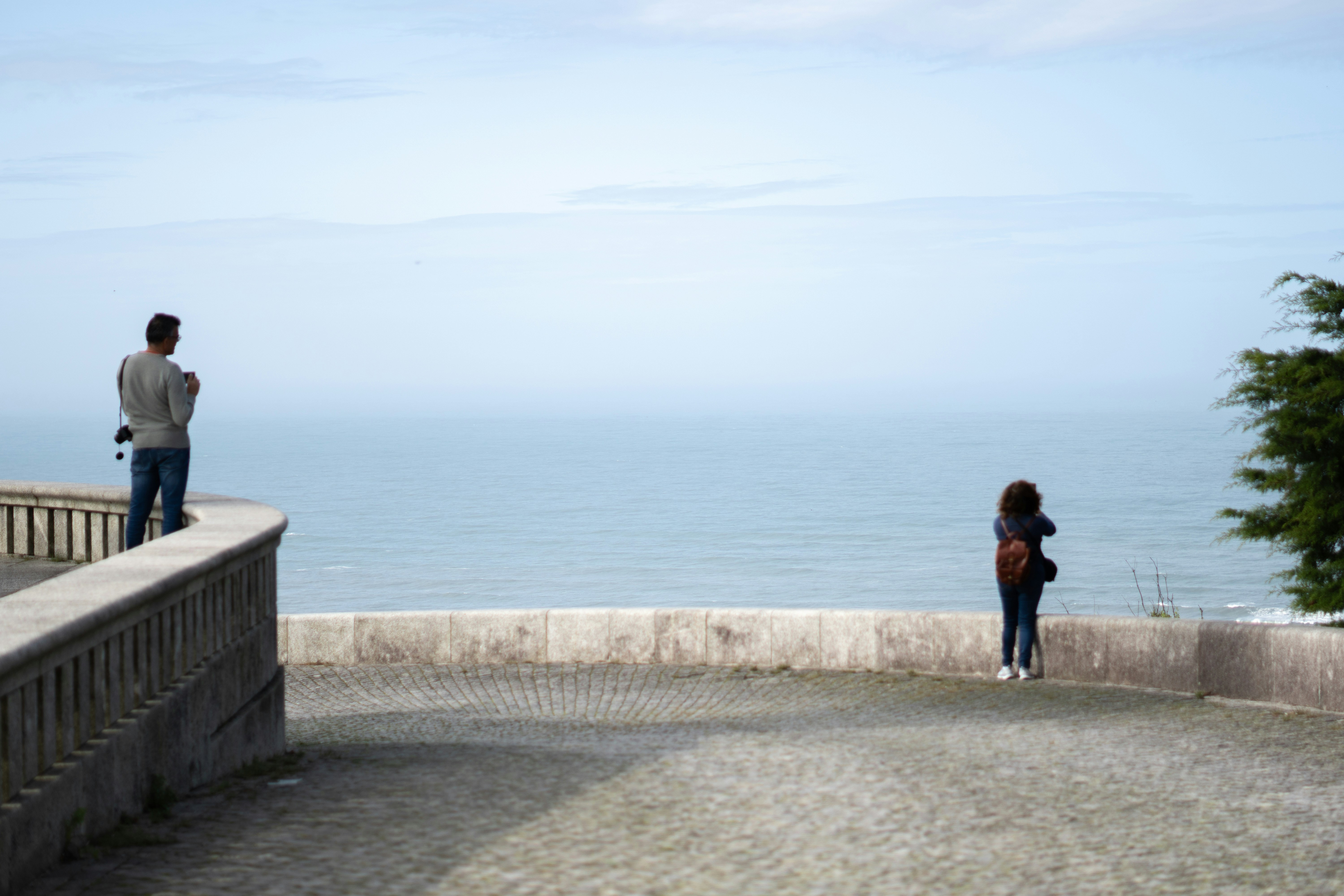 Two individuals stand apart on a stone wall overlooking a calm ocean under a cloudy sky.