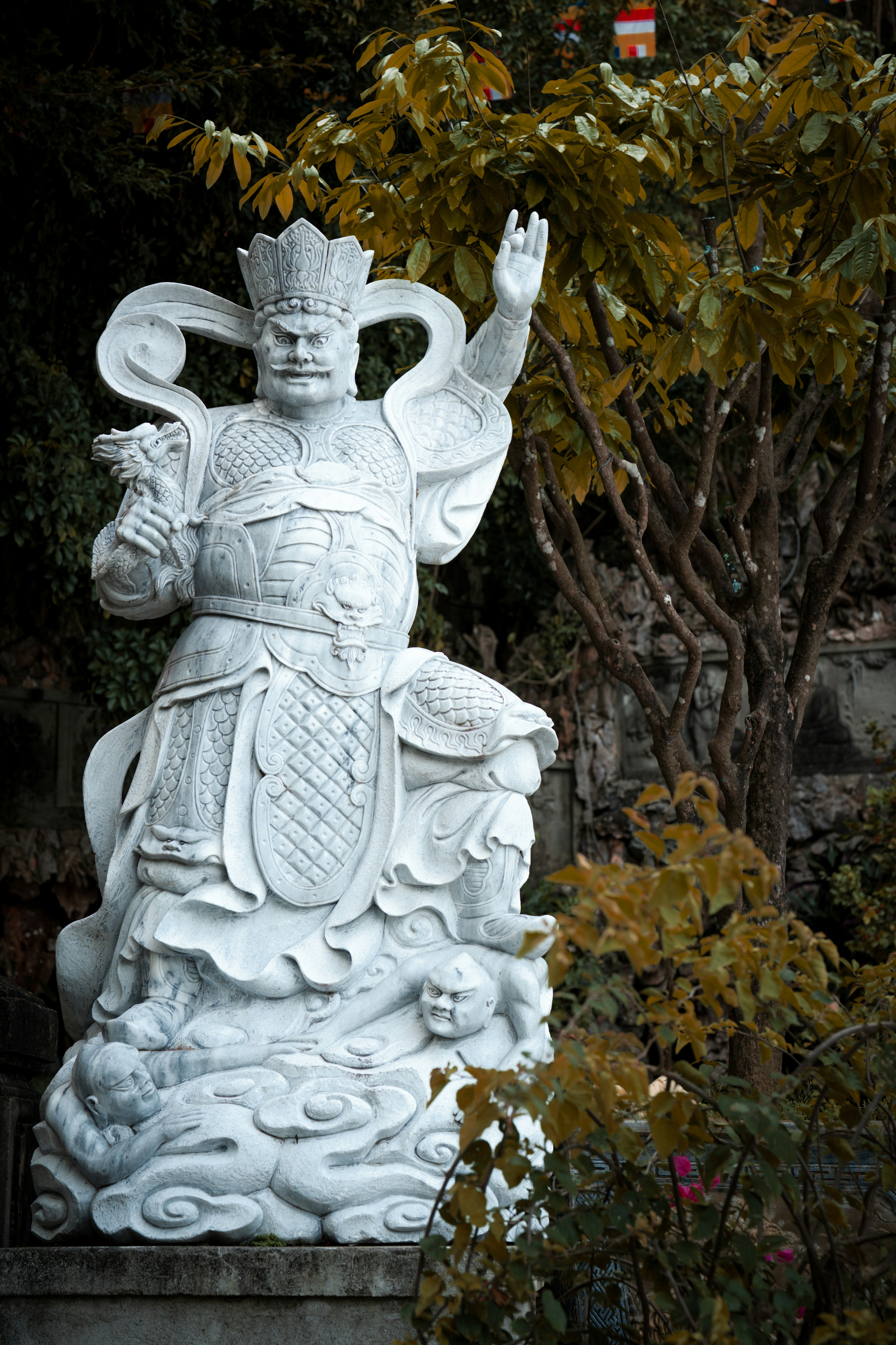 White marble statue of a warrior deity surrounded by trees with autumn foliage.