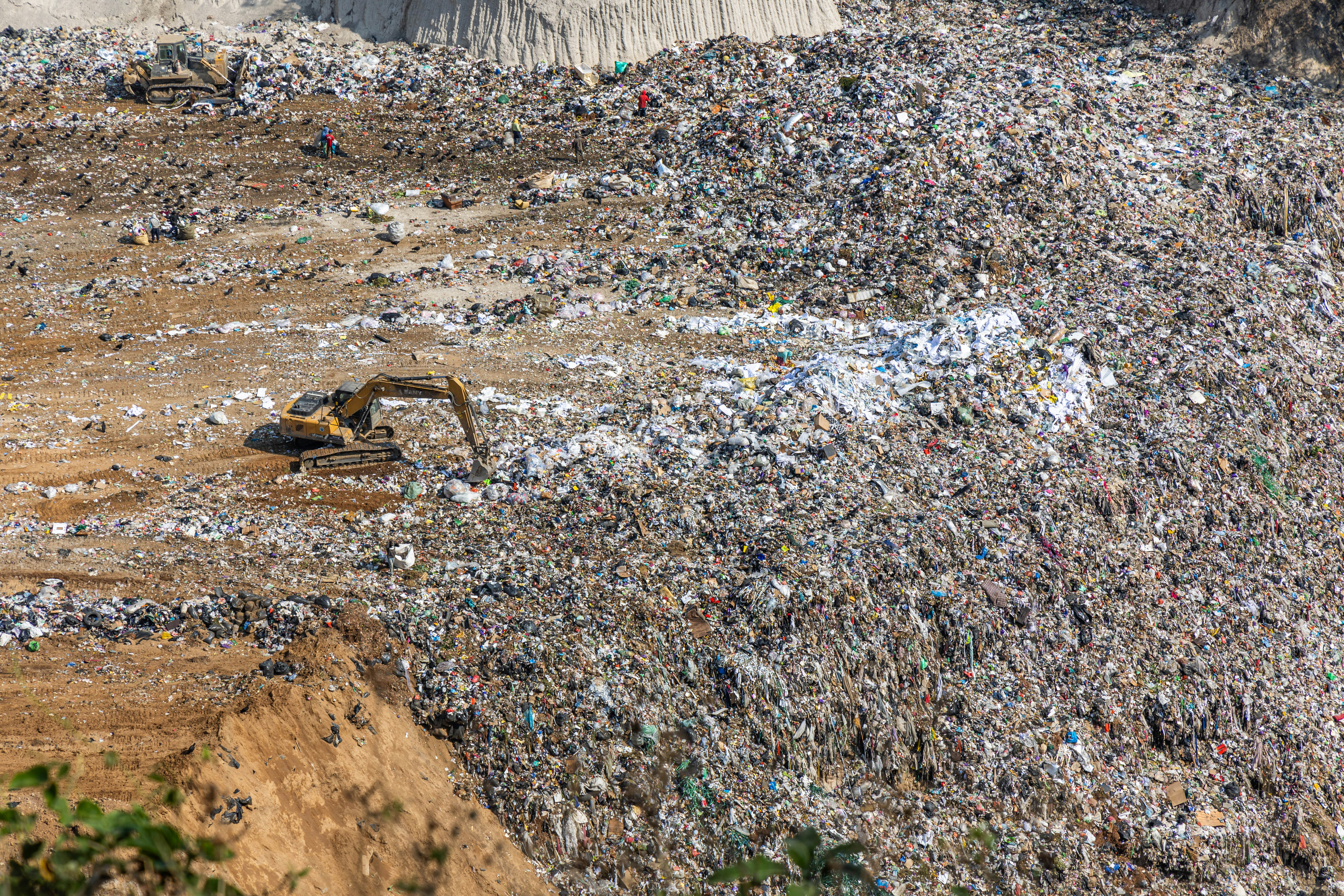 Excavators maneuver through extensive layers of debris at a vast landfill site, surrounded by steep earth walls.
