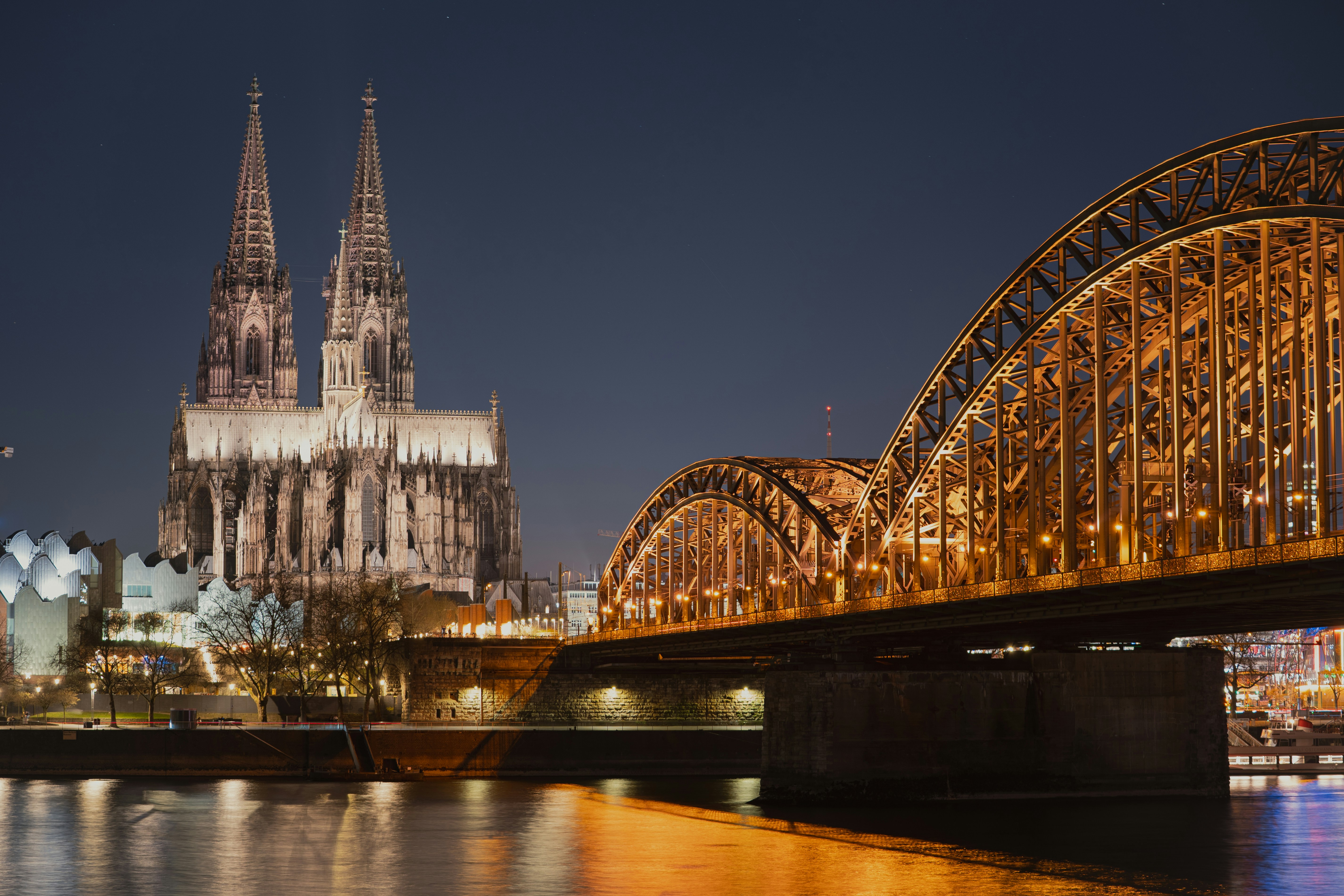 Cologne Cathedral and Hohenzollern Bridge illuminated at night, reflected in the calm waters of the Rhine River.