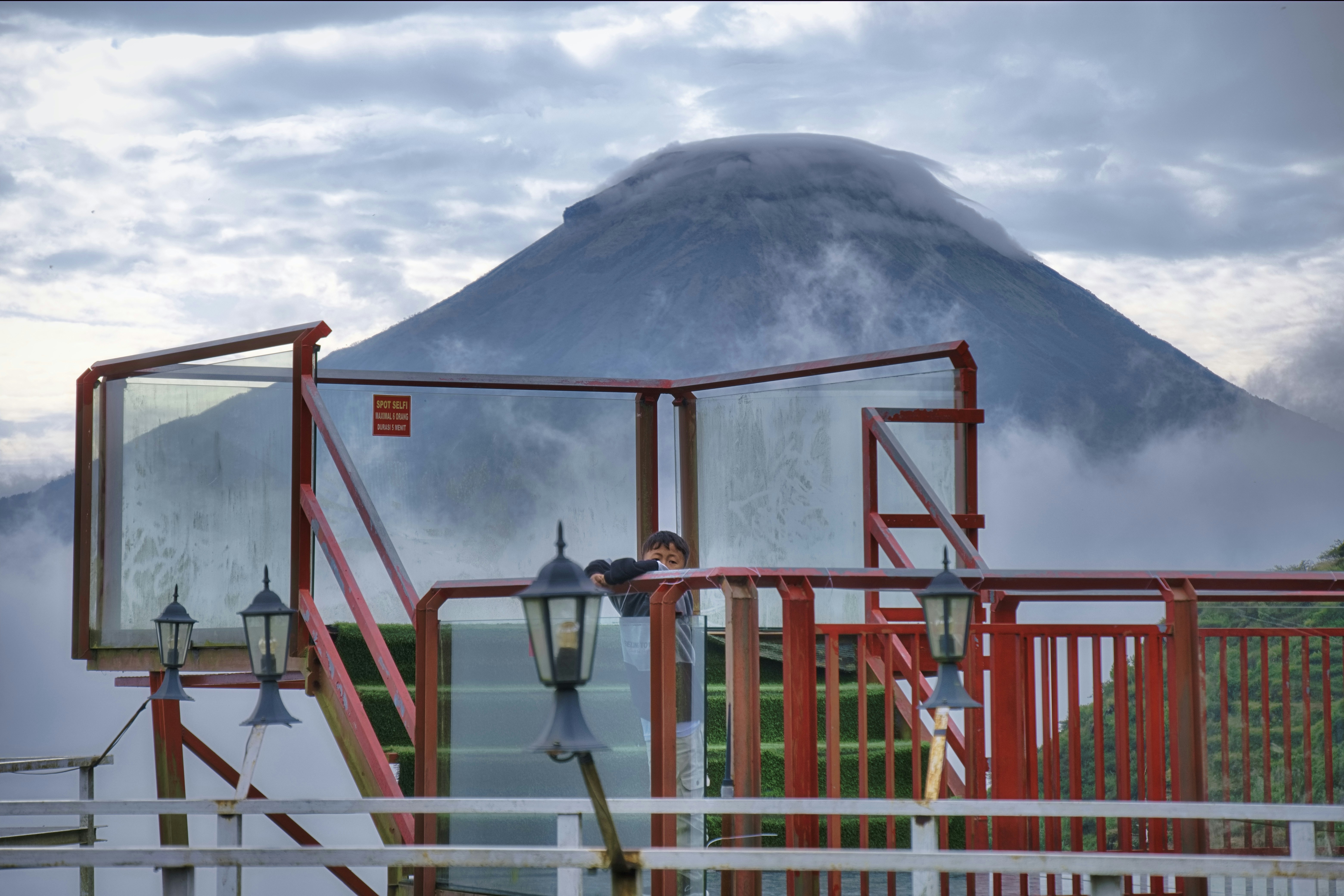 A breathtaking view of a mountain in the Dieng Plateau, partially covered in mist, creating a surreal atmosphere. A person leans on a red observation deck, adding a human element to the scene. The contrast between the structured platform and the wild, foggy landscape enhances the sense of depth and scale.
